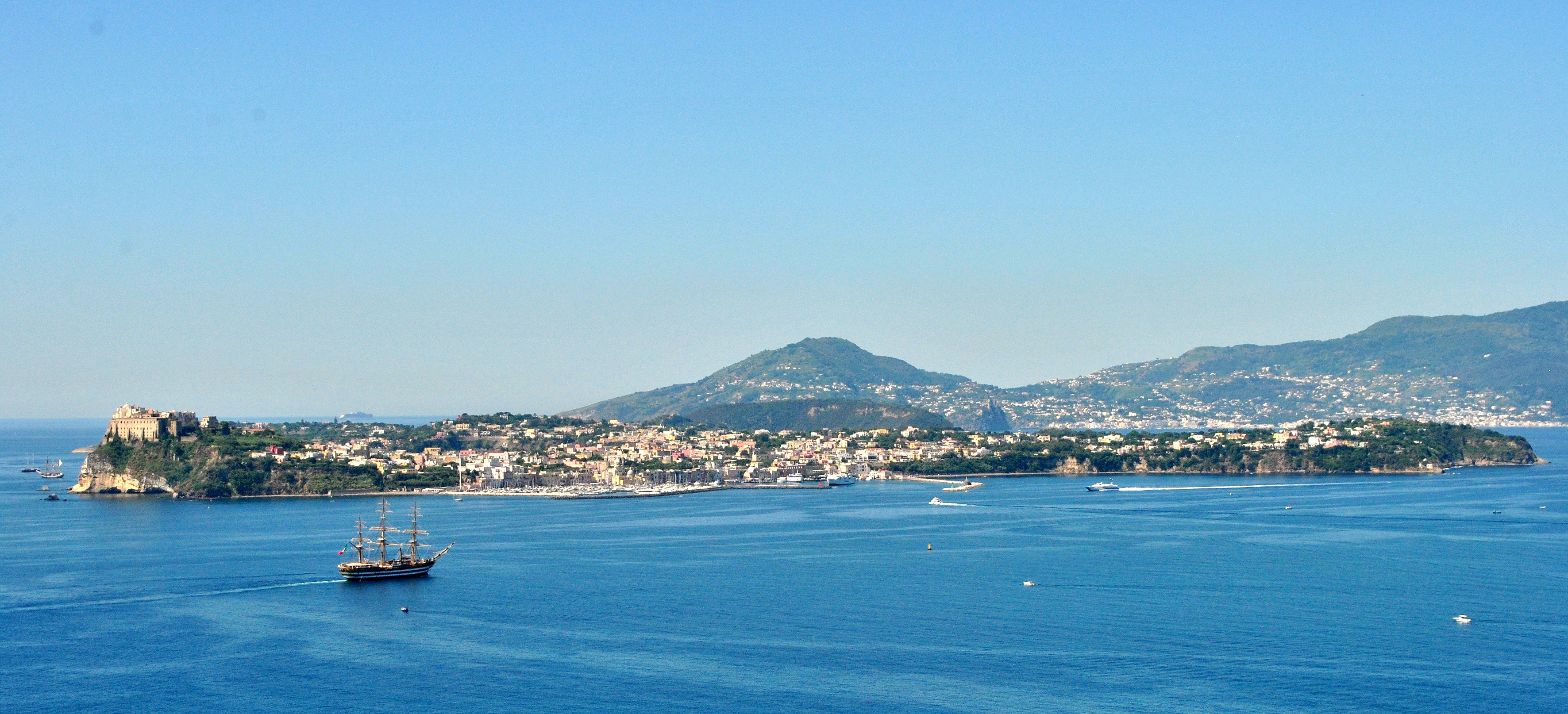Coastal town under clear blue skies with distant mountains and a sailing ship on calm waters.
