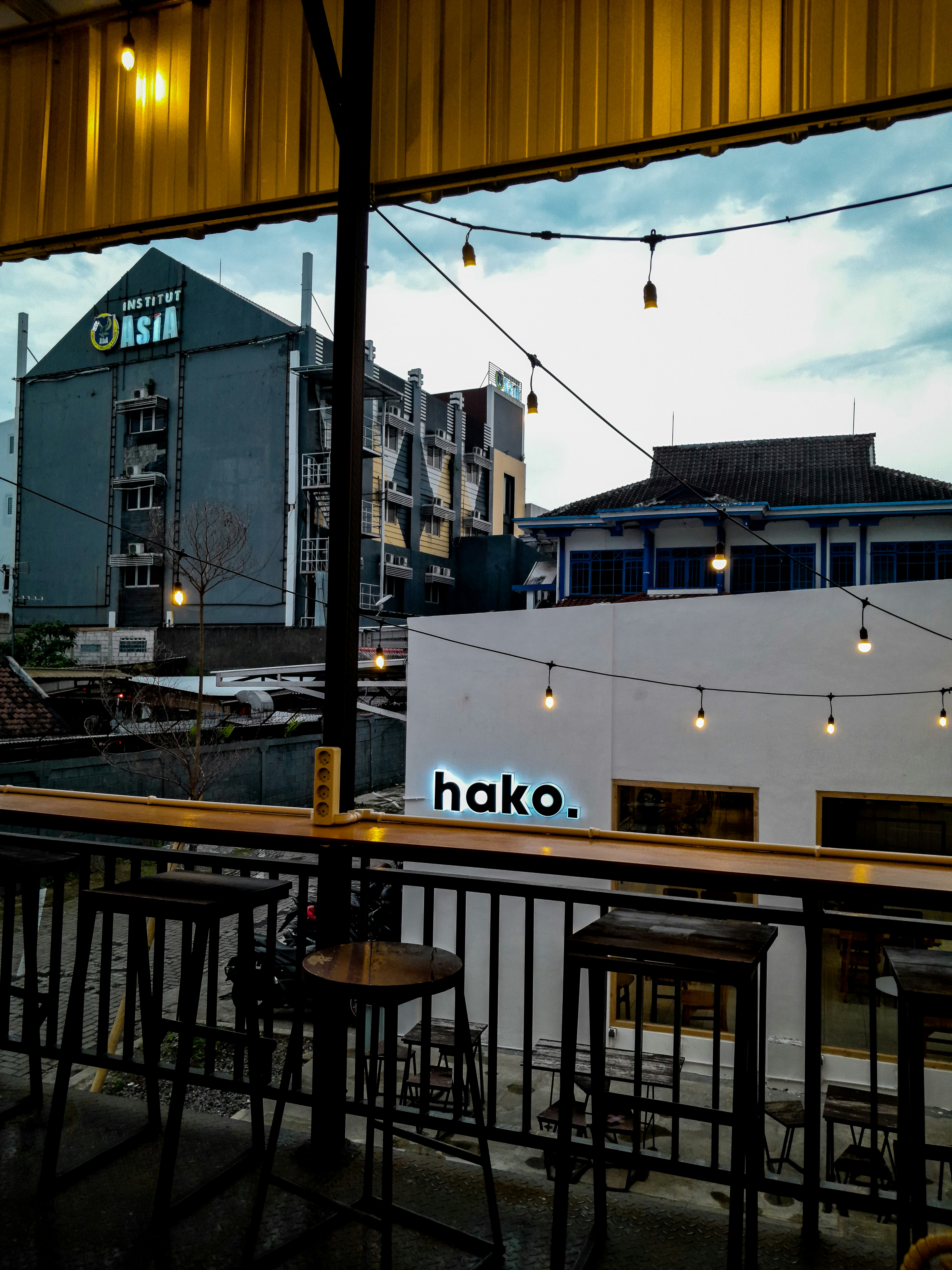Rooftop bar scene at dusk with string lights and a white wall bearing the illuminated 'hako.' sign. Urban buildings rise in the background behind railings and stools.