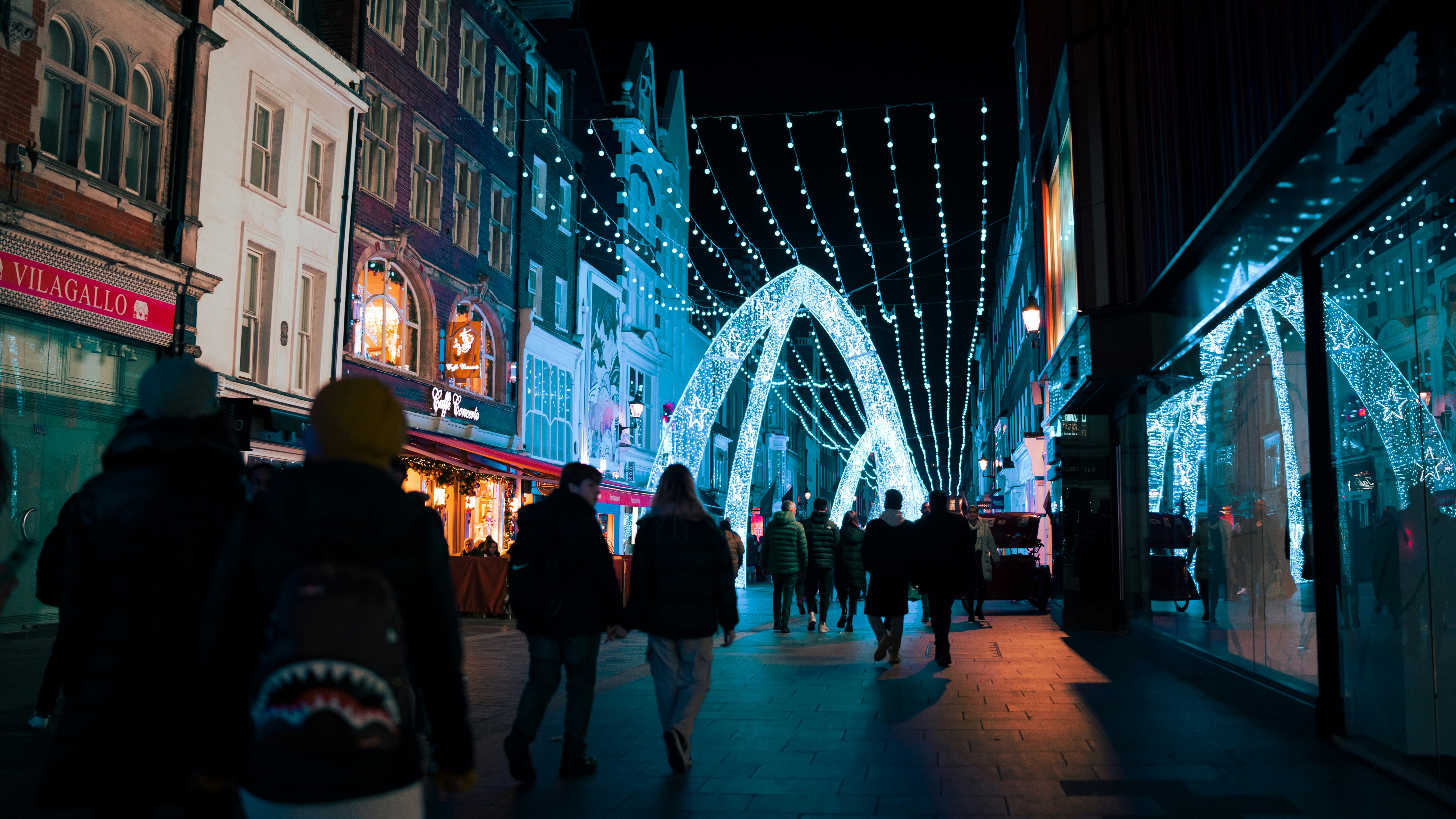 This captivating image showcases a bustling city street adorned with brilliant festive lights, creating an enchanting atmosphere. The composition leads the eye through an arch of glowing bulbs, with reflections in nearby windows enhancing the depth and vibrancy of the scene. The cool blue and white tones contrast beautifully with the warm hues from shop windows, capturing the lively essence of a winter evening.