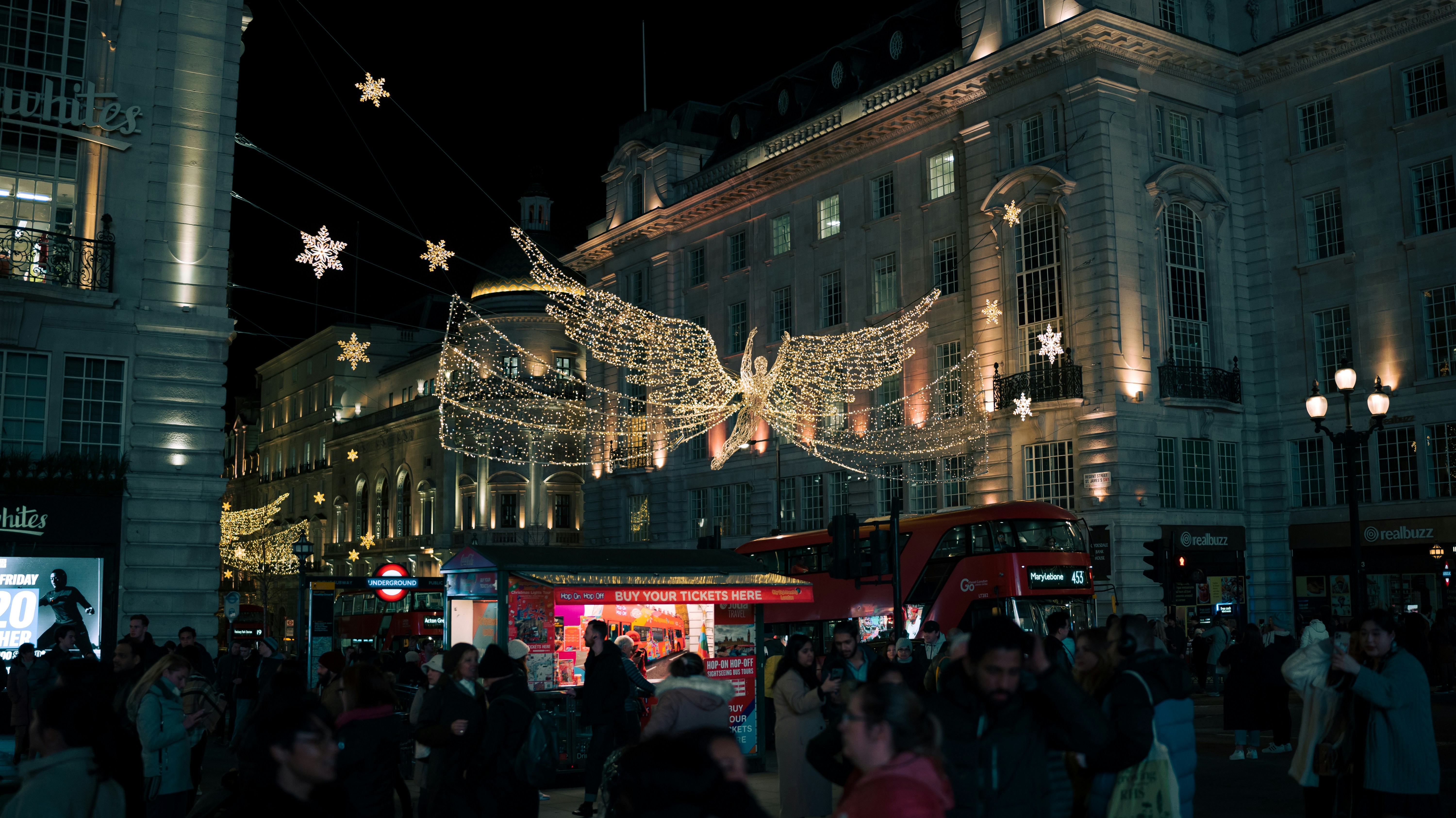 This captivating image captures the vibrant energy of a city street adorned with festive lights. A magnificent angel made of twinkling golden lights hovers above the bustling crowd, casting an enchanting glow over the scene. The warm illumination contrasts beautifully with the cool night sky, highlighting the elegant architecture of the surrounding buildings and creating a lively, celebratory atmosphere.