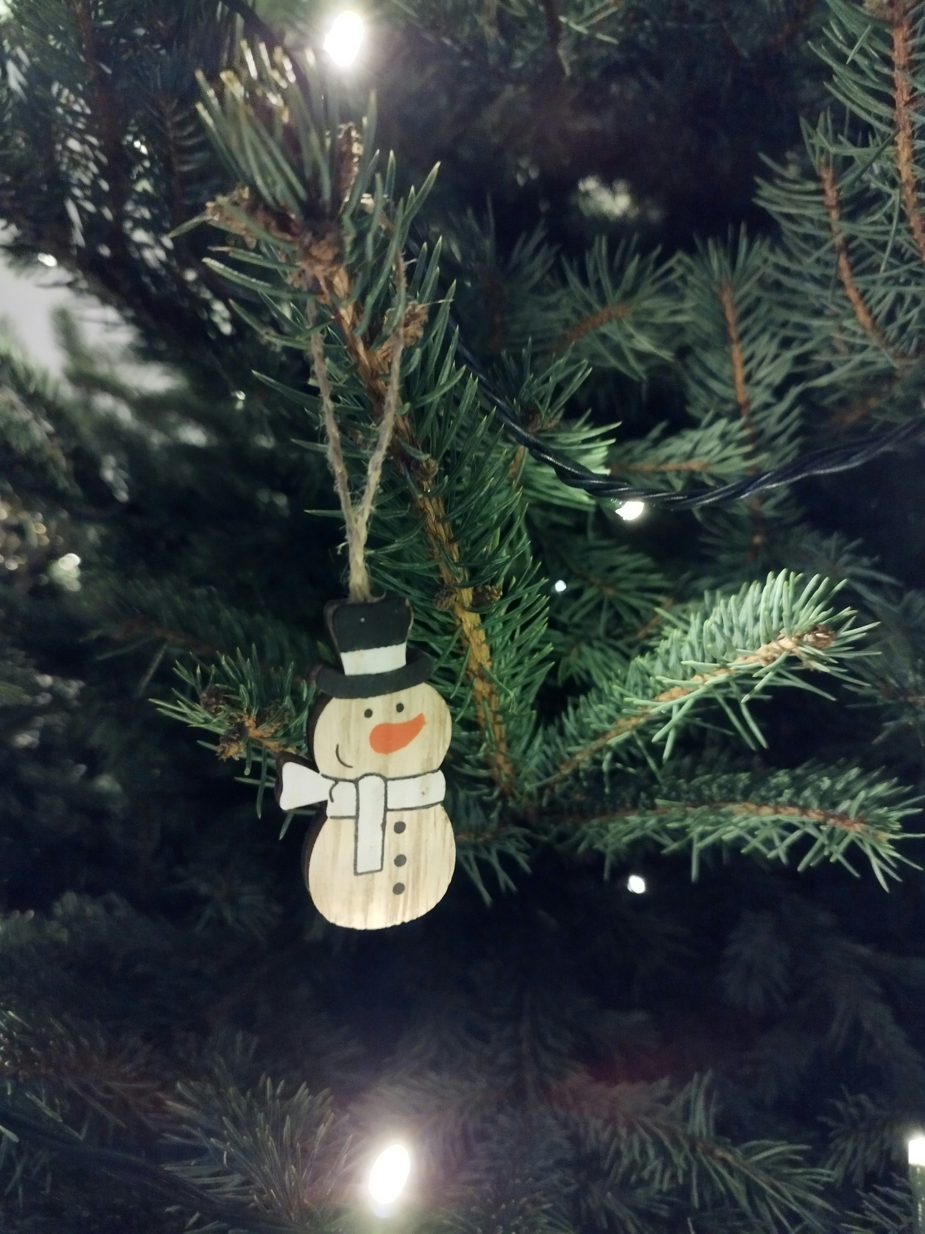 Close-up photo of a wooden snowman ornament hanging from a pine tree, accented by string lights.