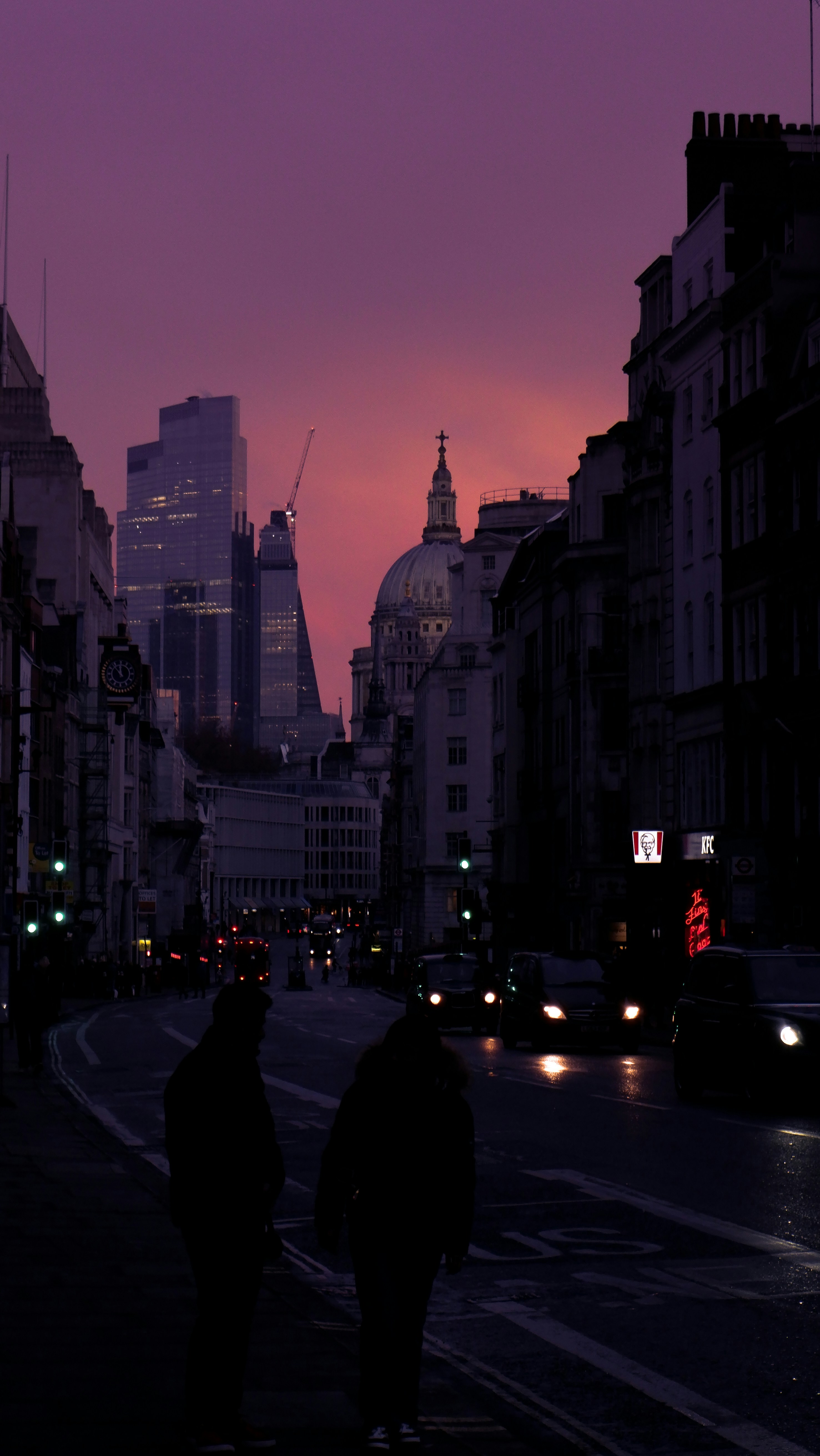 Silhouetted figures stand on a city street as skyscrapers loom in the background, illuminated by a vibrant twilight sky.