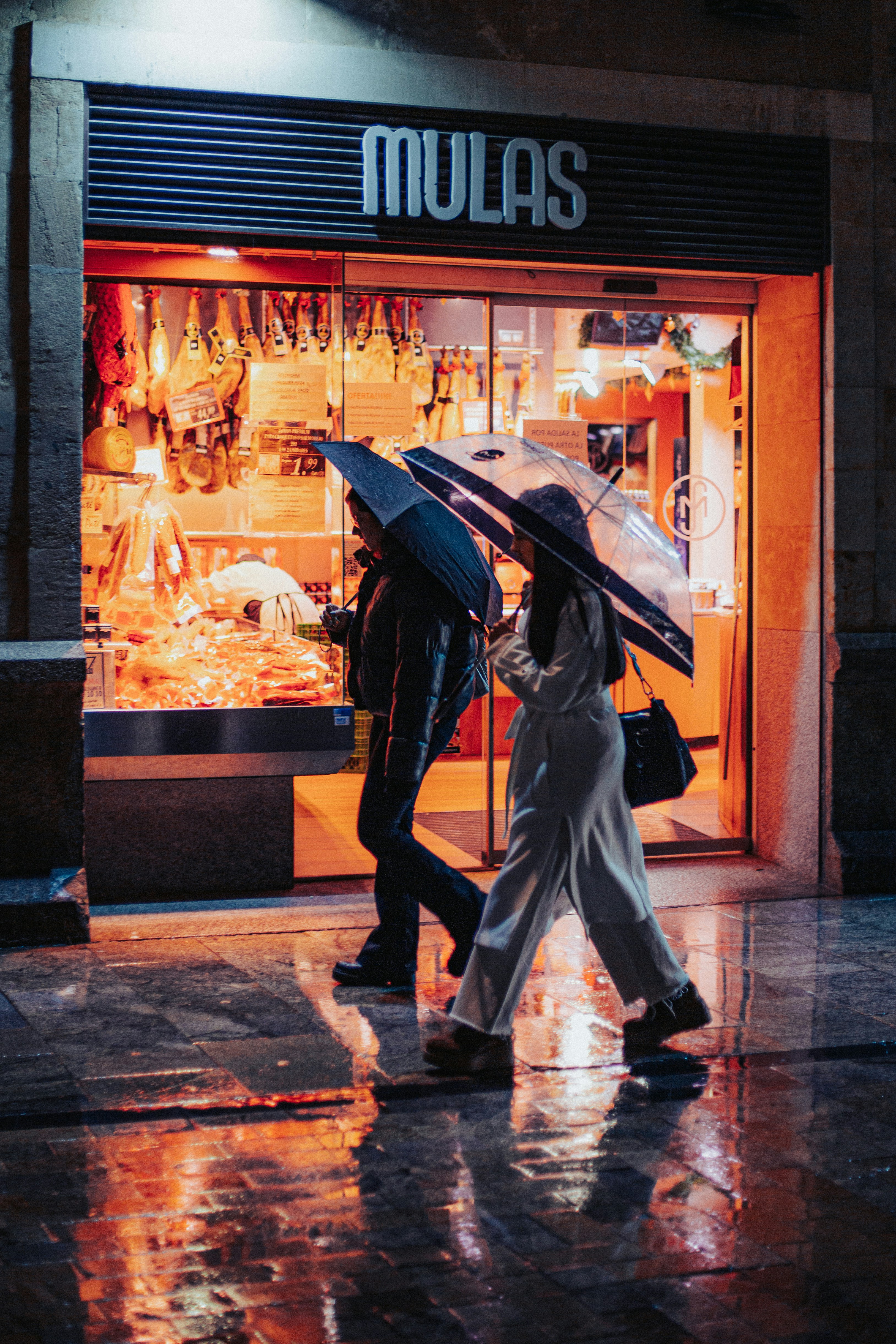 Two people with umbrellas walk past an illuminated shop window on a rainy night.