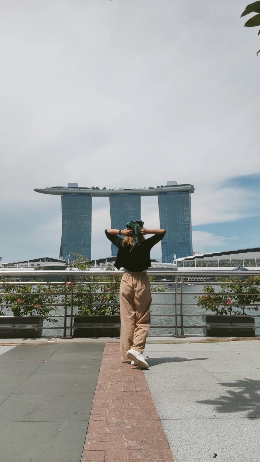 Person standing with hands on head facing Marina Bay Sands hotel and skyline from a waterfront promenade.