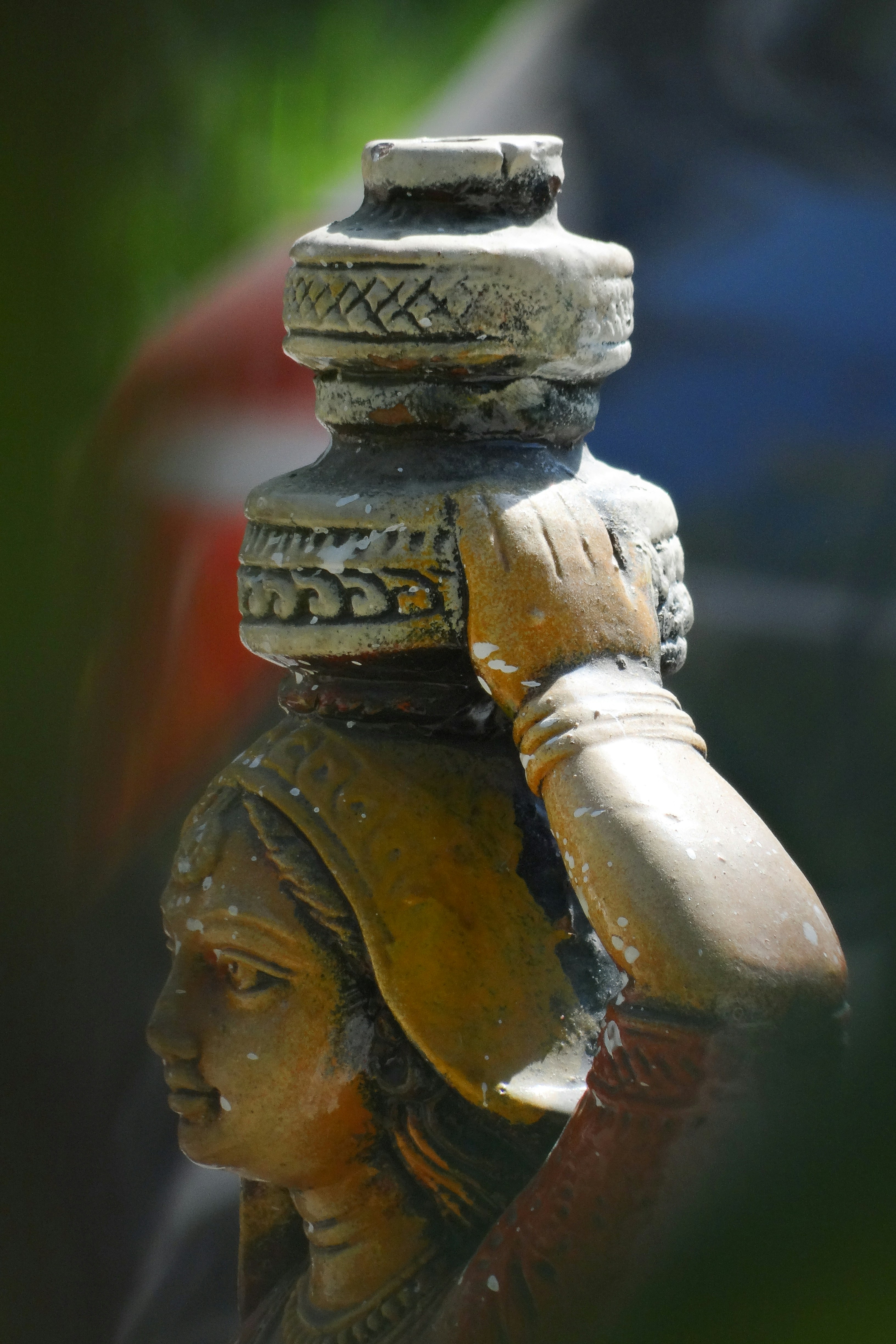 Close-up of a weathered sculpture depicting a figure balancing an ornate urn on the head, with a softly blurred garden backdrop.