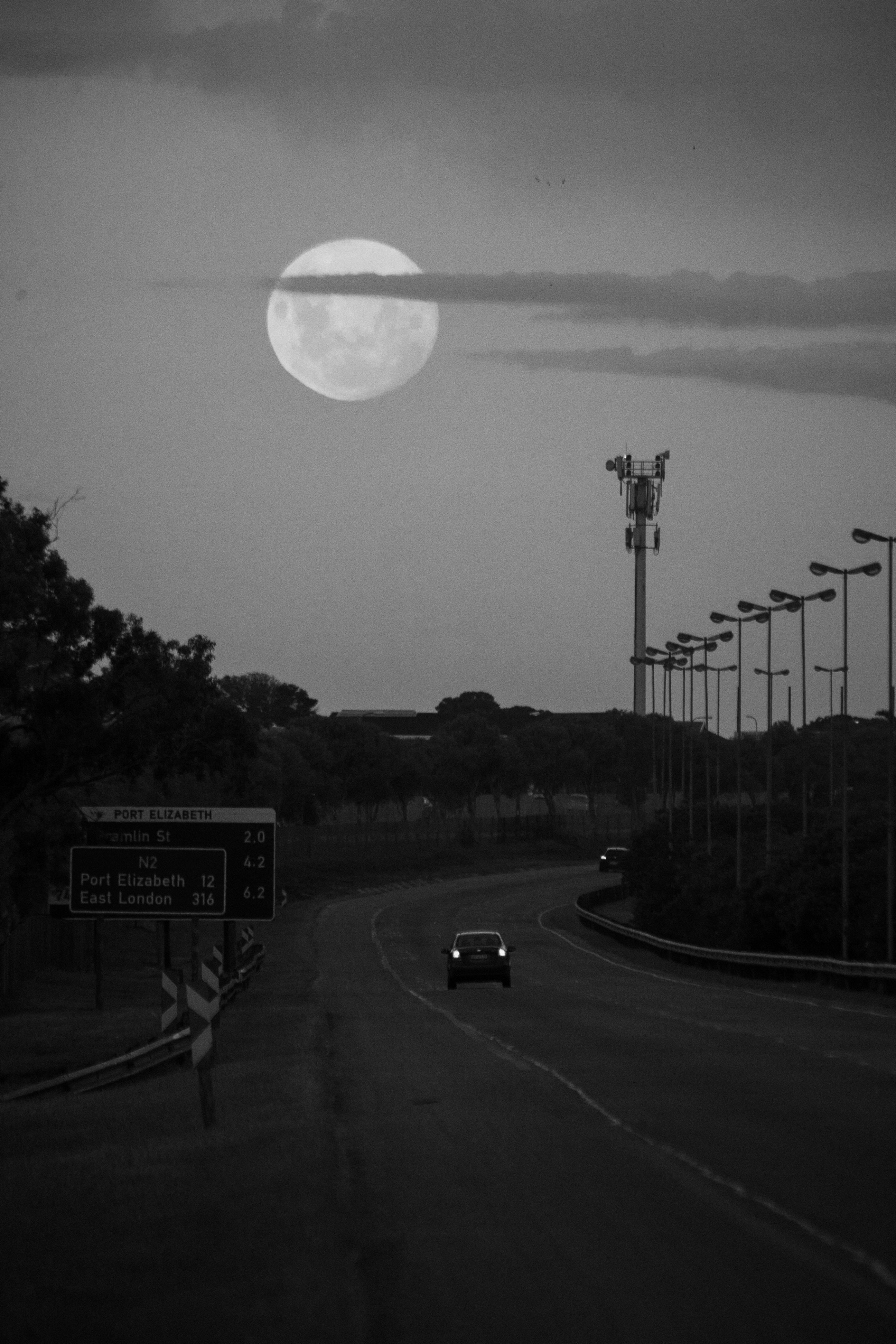 A solitary car travels along a winding road under the watchful gaze of a large moon partially obscured by clouds, creating an atmospheric scene.