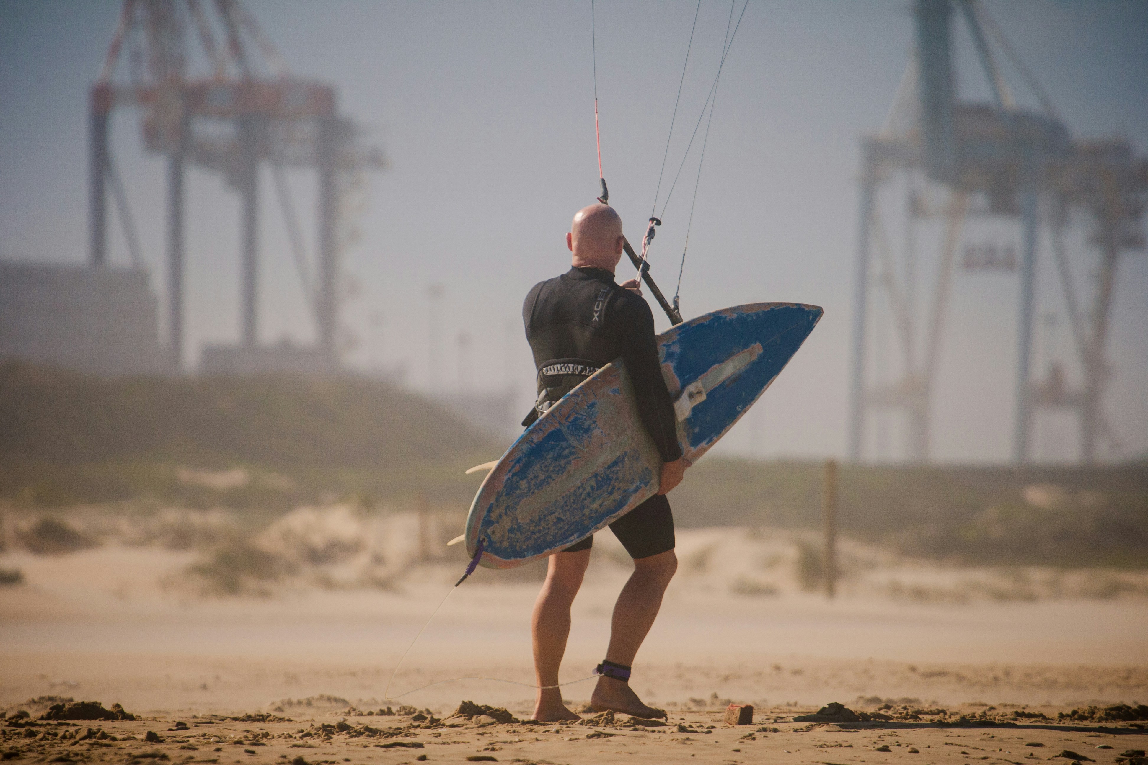 Solitary Surfer with Weathered Blue Board on a Beach Framed by Industrial Cranes