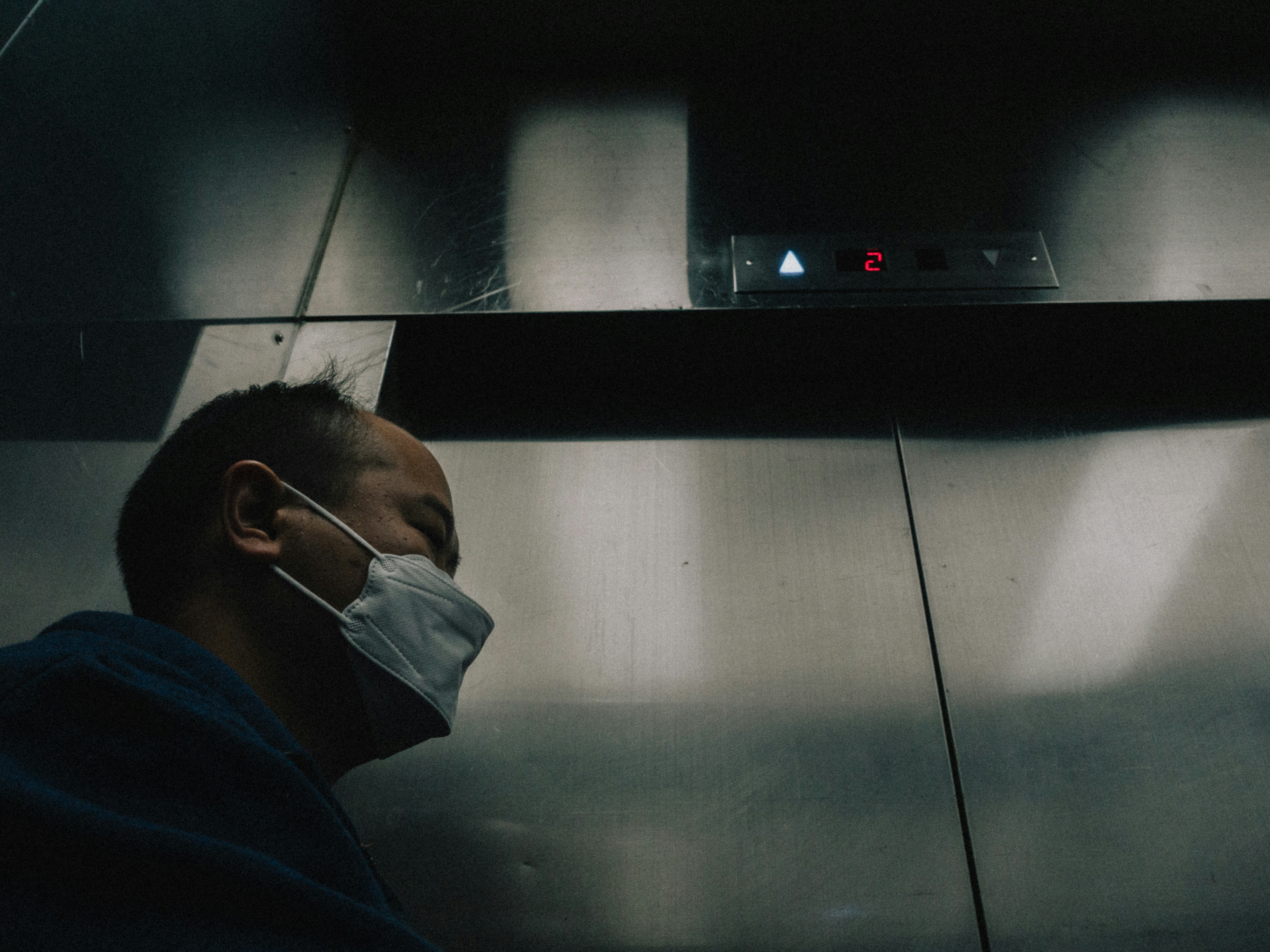 The image captures a solitary moment inside an elevator, focusing on a person wearing a face mask, enveloped in the cool metallic reflection of the elevator walls. The subdued lighting casts a moody ambiance, with soft shadows and a hint of the elevator's digital panel displaying the number '2'. The muted tones of grey and blue create a somber yet contemplative atmosphere, making the composition visually striking and evocative of introspection.