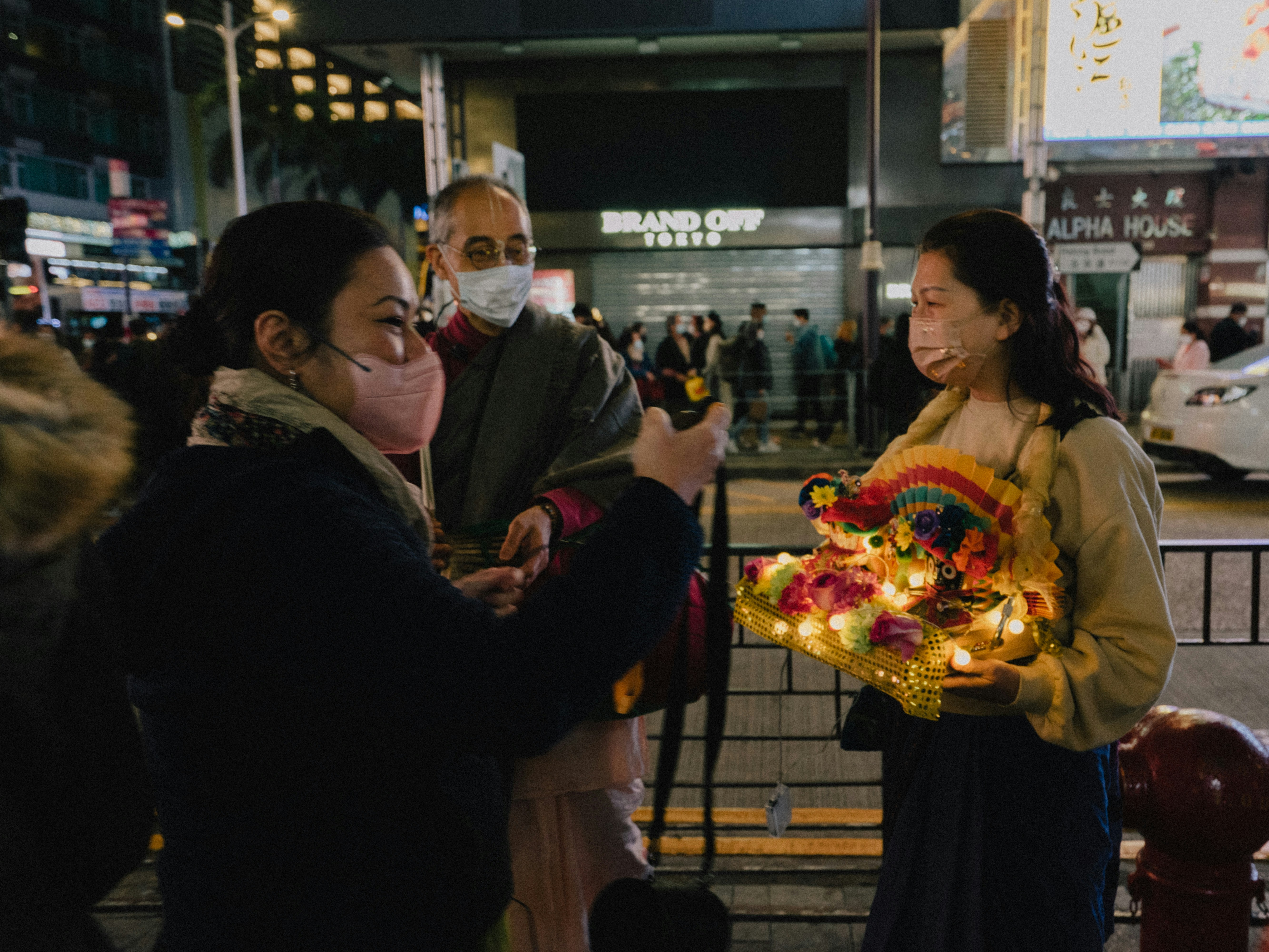 In this lively street scene, a woman holding a brightly lit, colorful display engages in conversation with a passerby, creating a captivating focal point amidst the urban backdrop. The image captures the contrast between the vibrant yellows, reds, and blues of her display against the subdued, moody lighting of the evening city. The ambient glow and bustling atmosphere evoke a sense of community and cultural richness, making the image visually striking and immersive.
