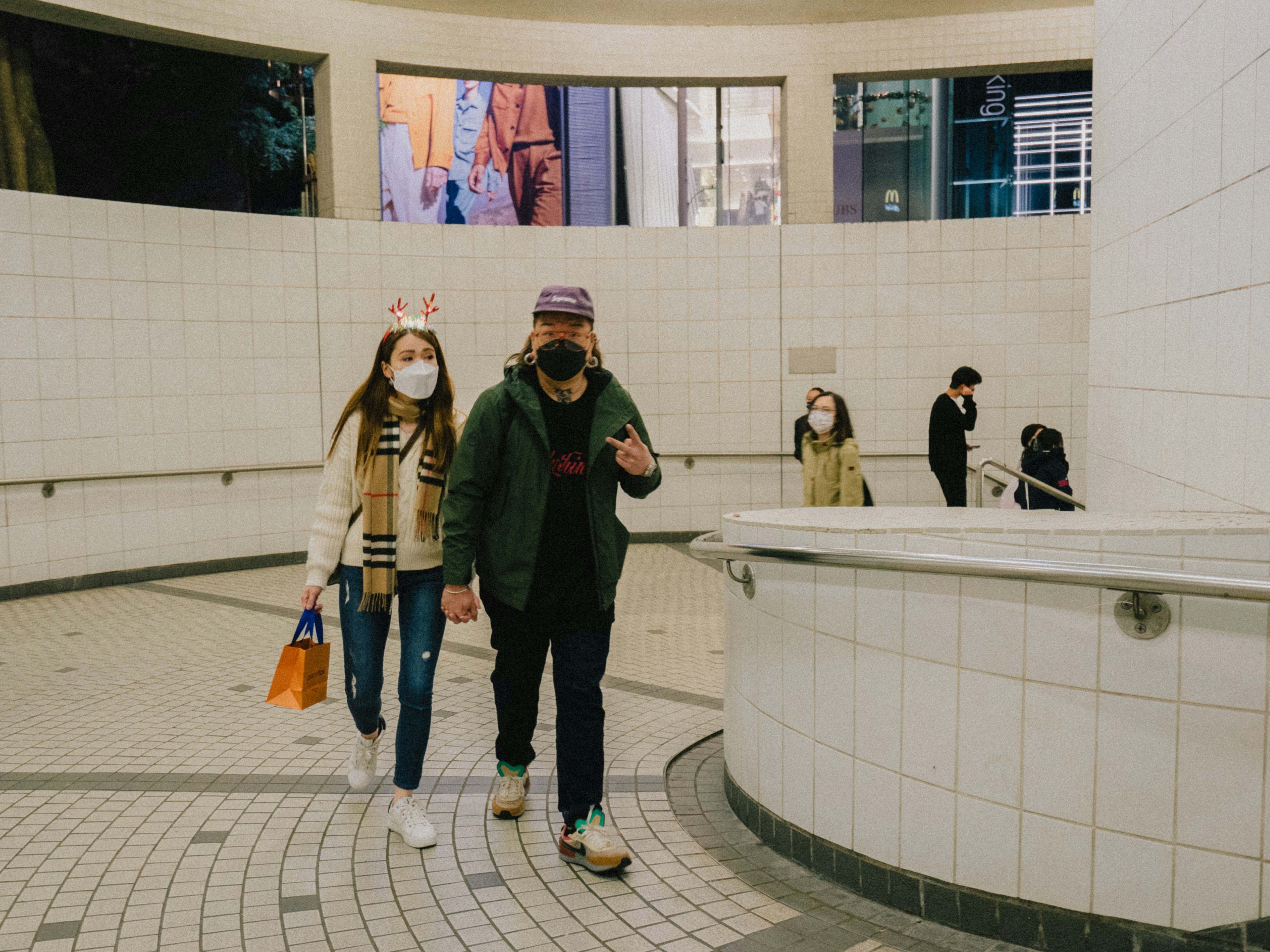 This candid shot captures a couple walking hand-in-hand through an urban underground passage, both adorned in winter attire and festive reindeer antlers. The composition is framed by the clean, geometric lines of the tiled walls, with a soft, diffused lighting that creates a cozy atmosphere. The contrast between the warm clothing and the cool tones of the surroundings highlights the lively, yet serene essence of city life during the holiday season.