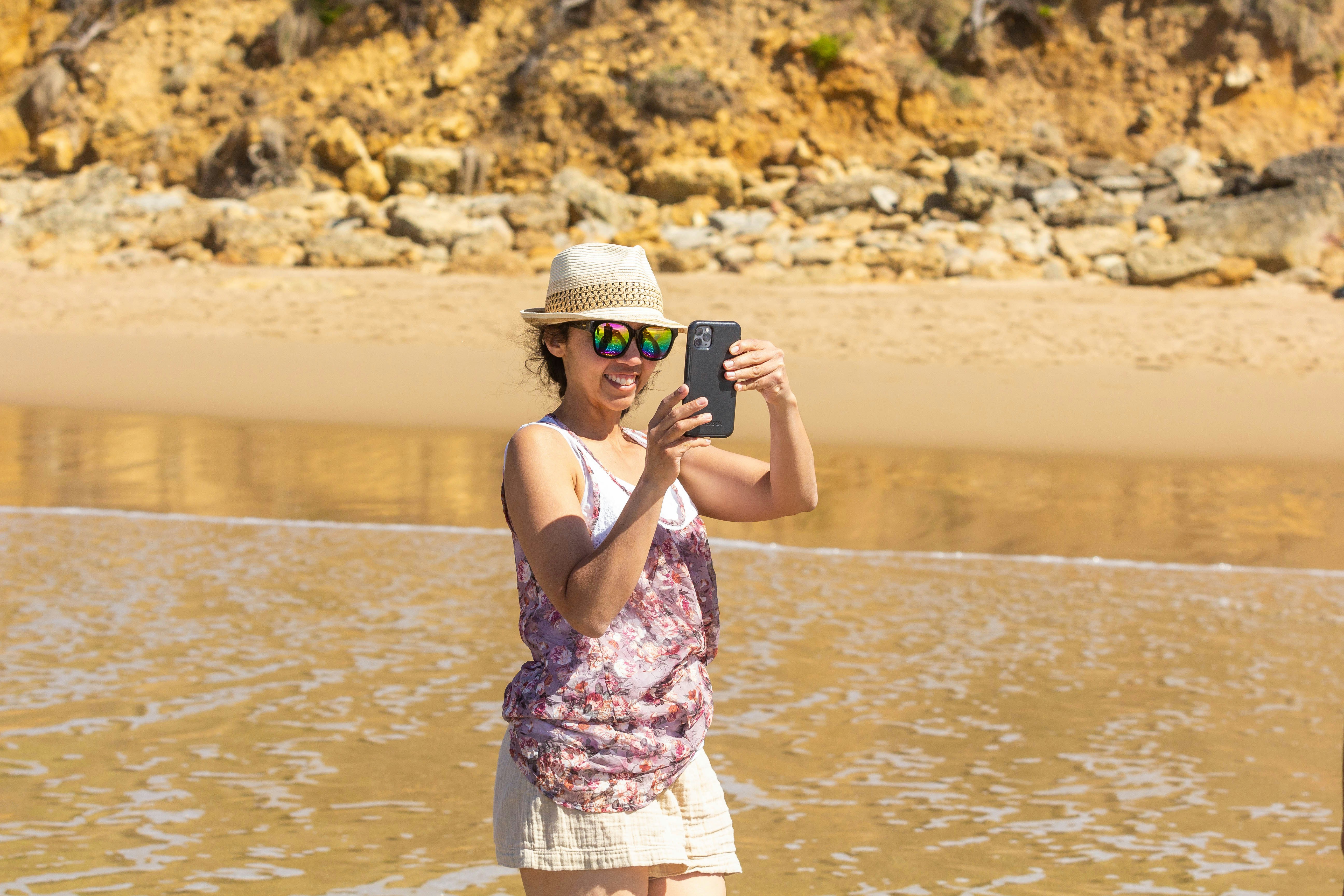 Selfie at the Shore: Straw Hat, Golden Beach, and a Rocky Backdrop