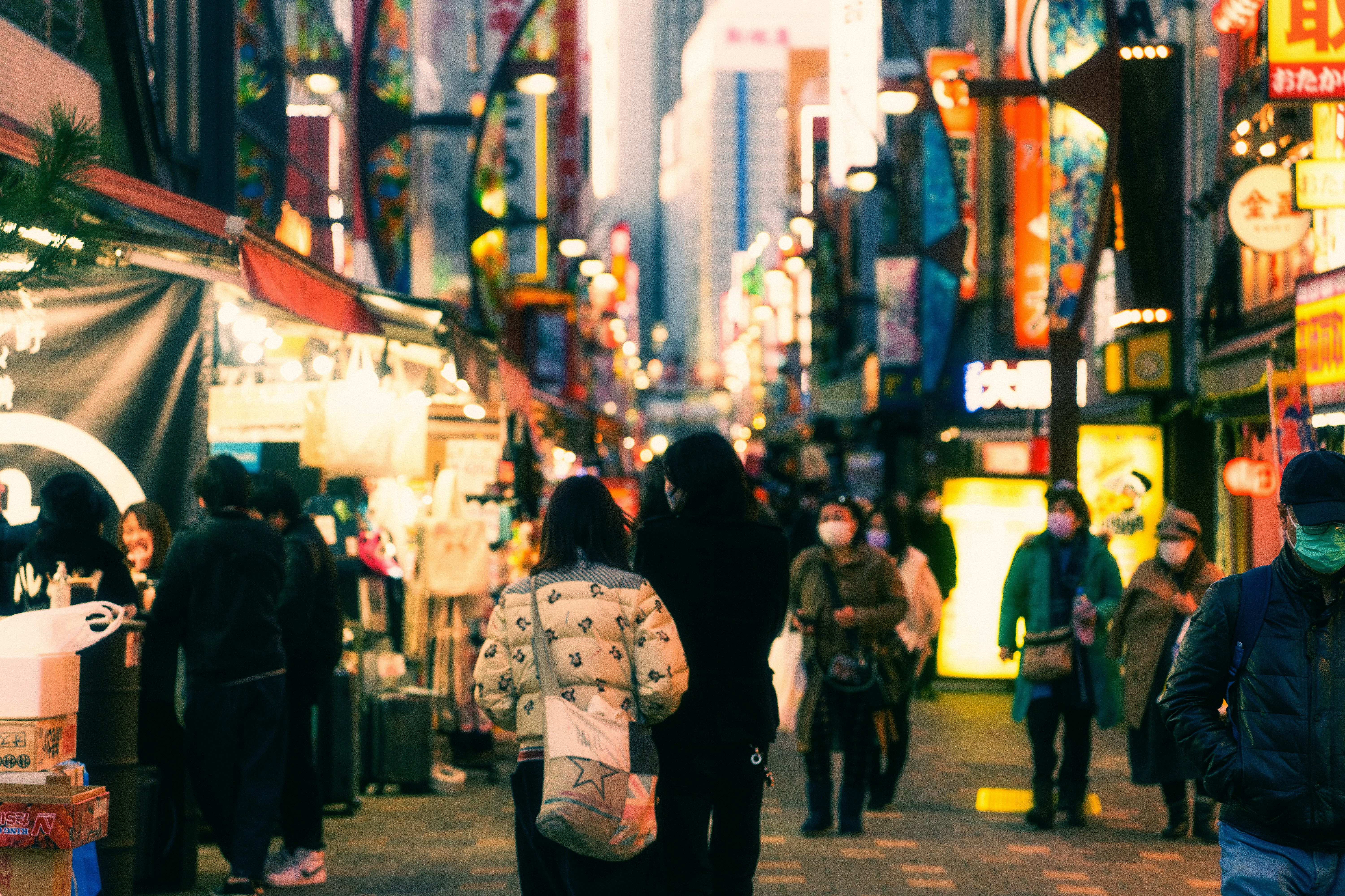 People walk through a bustling, neon-lit street market at night, surrounded by colorful signs and storefronts.