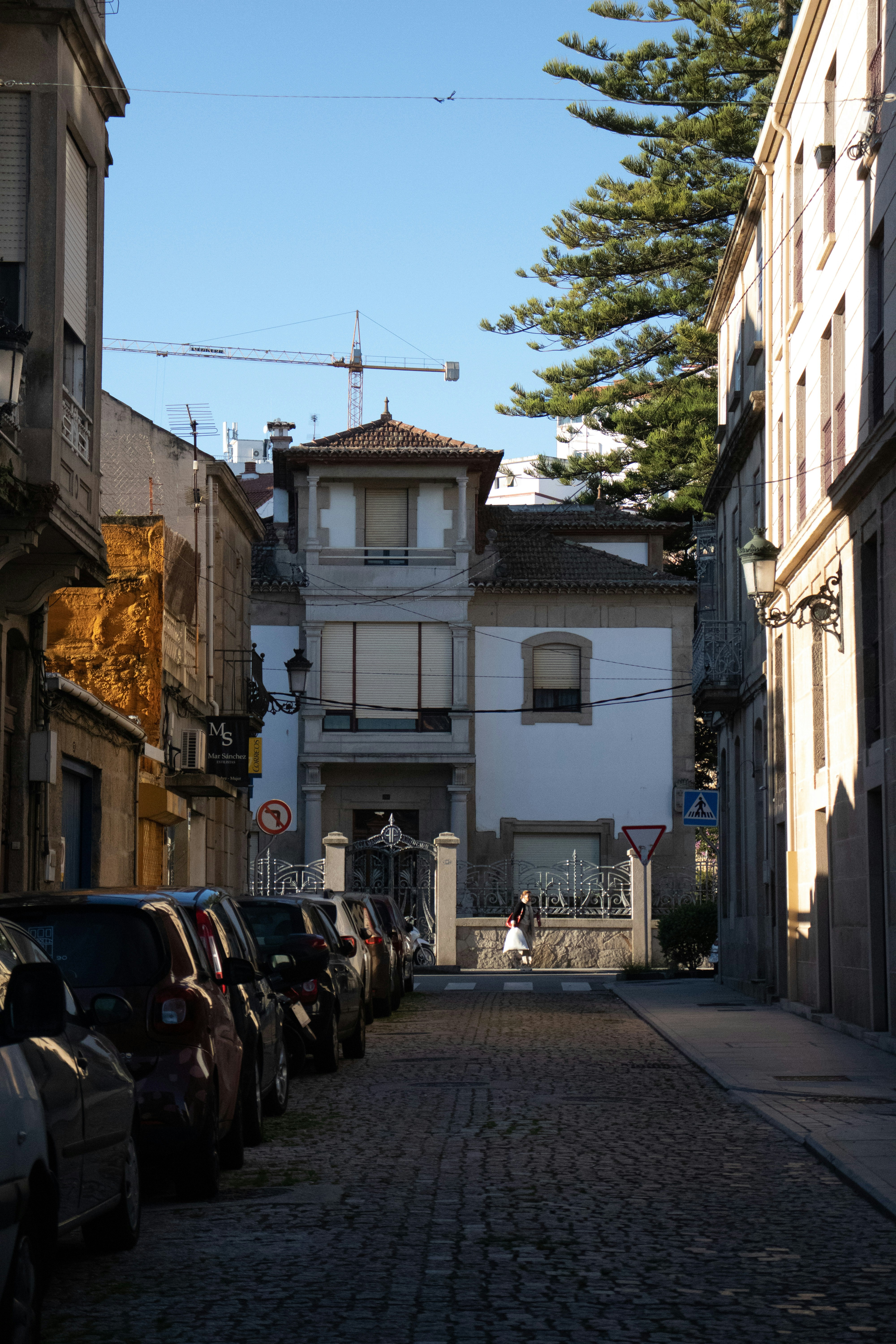Charming street scene featuring vintage architecture and parked cars lining a cobblestone road, with a person walking towards a gated entrance. 