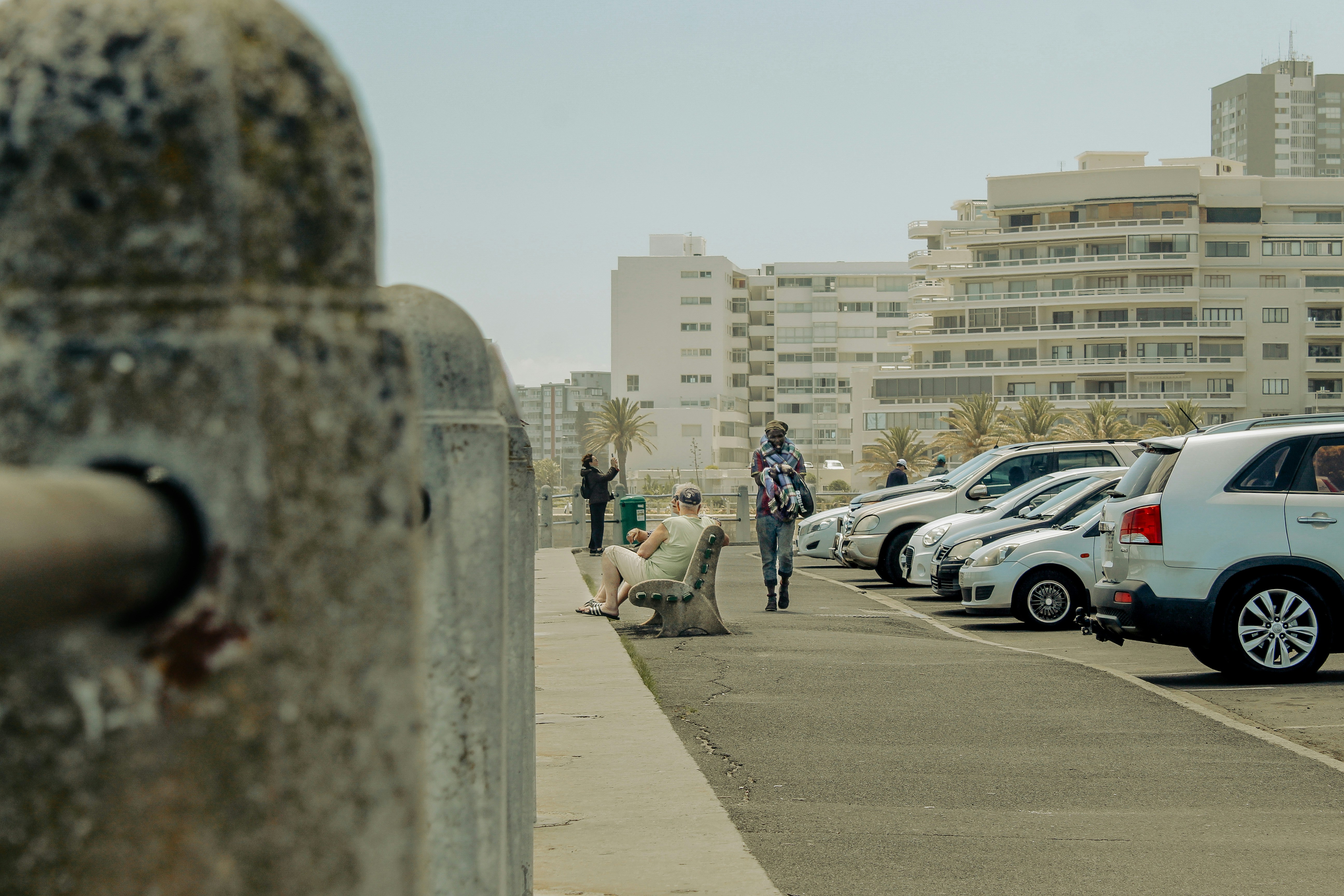 A relaxed scene at a coastal parking area, featuring individuals enjoying the sunny day amidst parked cars and modern buildings.