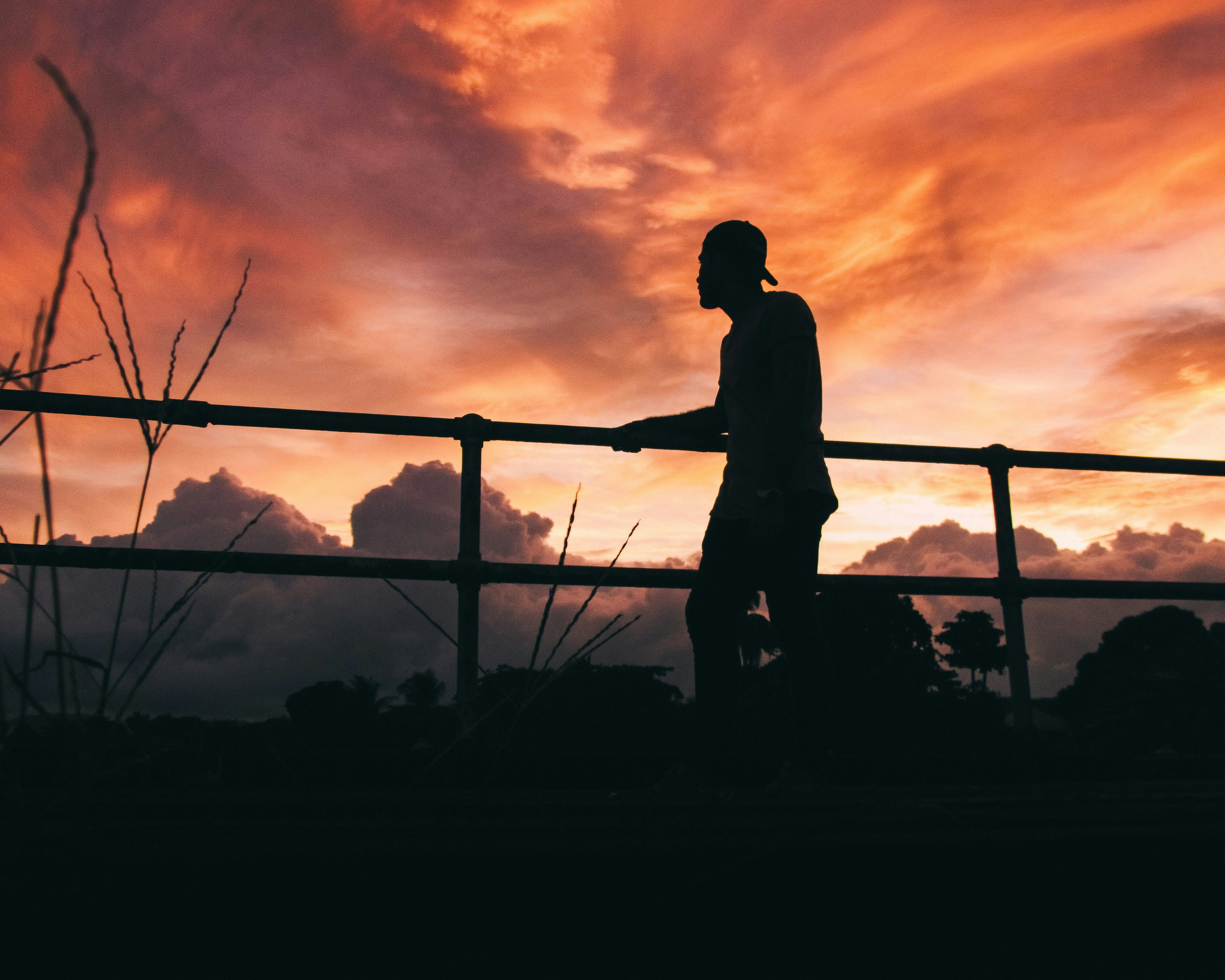Silhouette of a person leaning on a railing as vibrant clouds fill the sky at sunset.