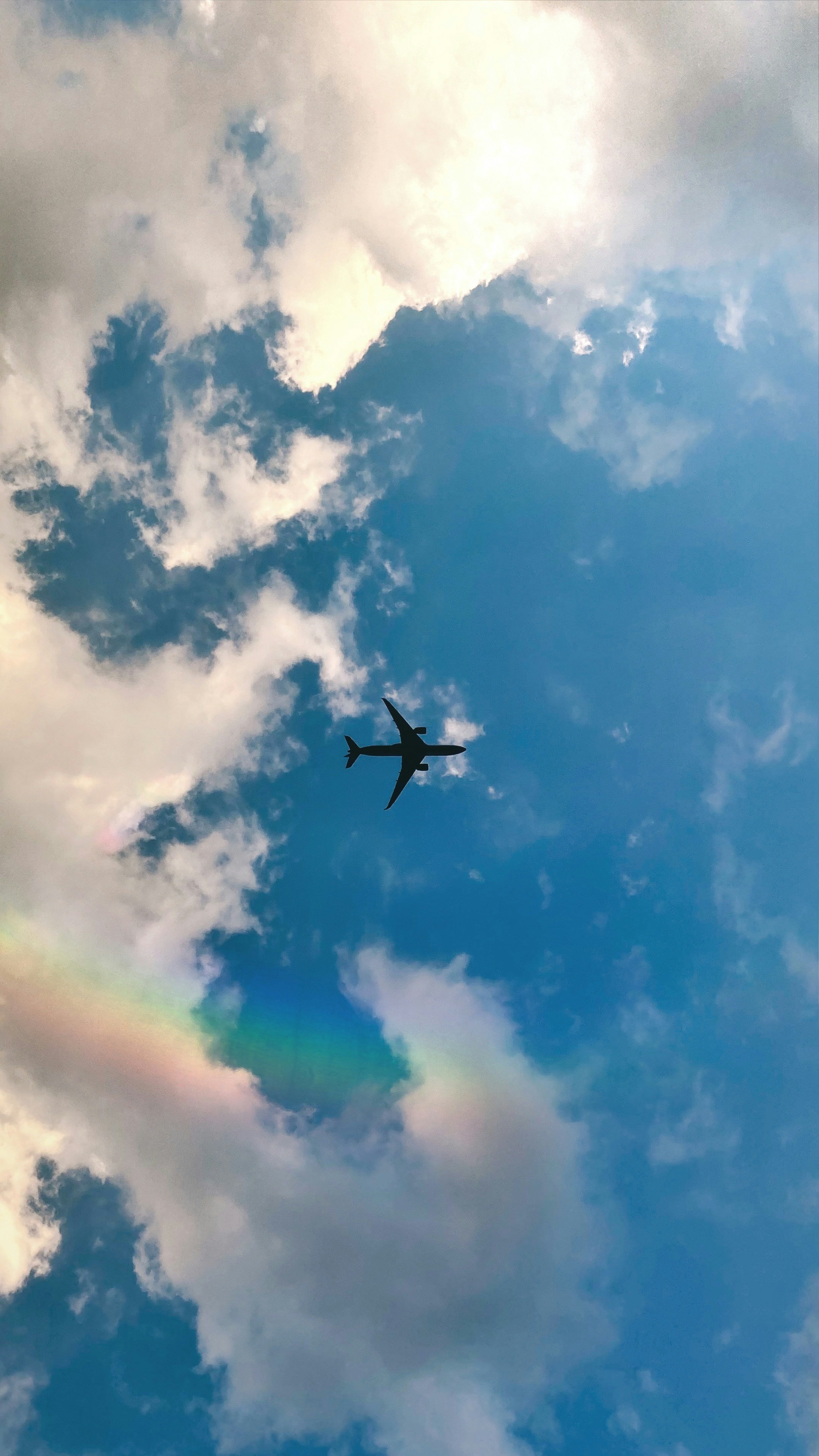 Airplane gliding beneath a vibrant sky adorned with clouds and a subtle rainbow arc.