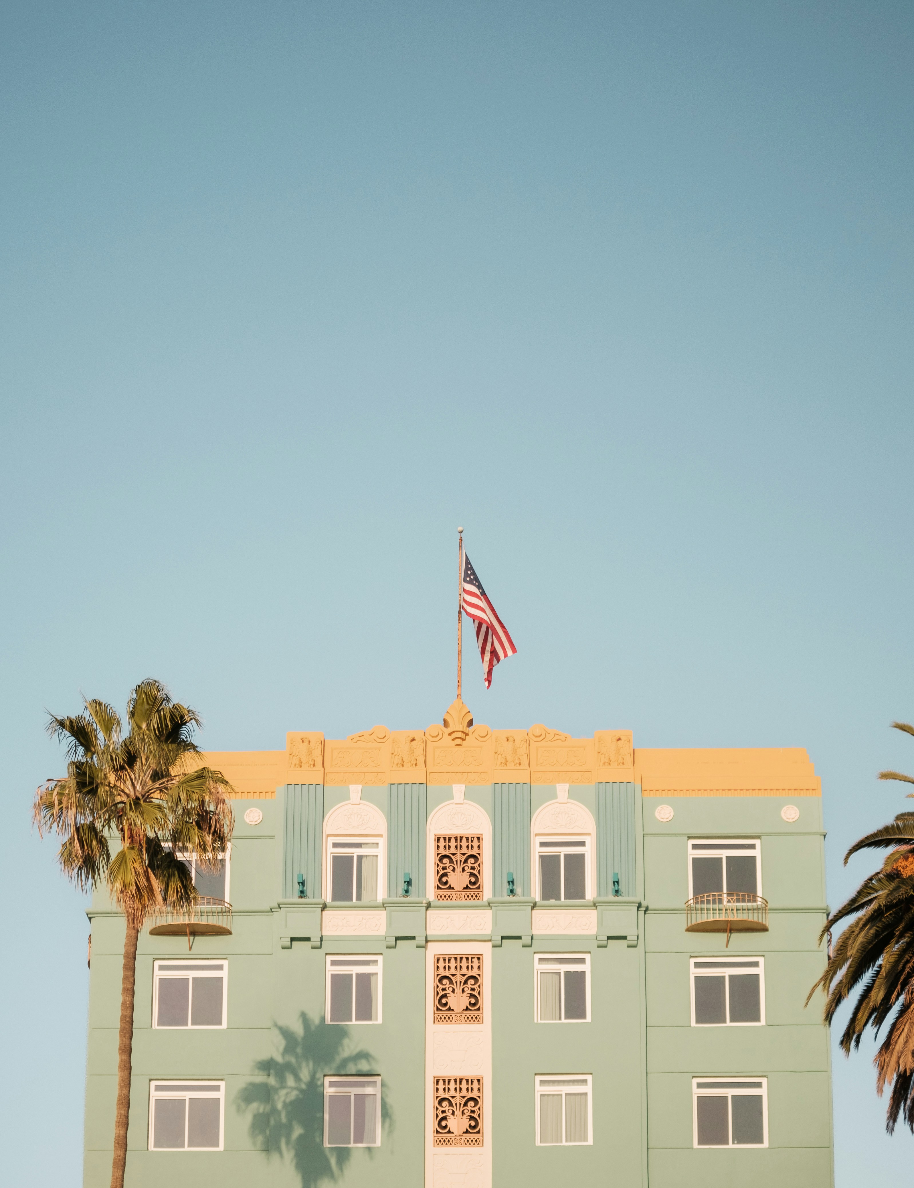 Flag Over a Seafoam Hotel: Palm-Framed Pastel Facade Under a Clear Sky
