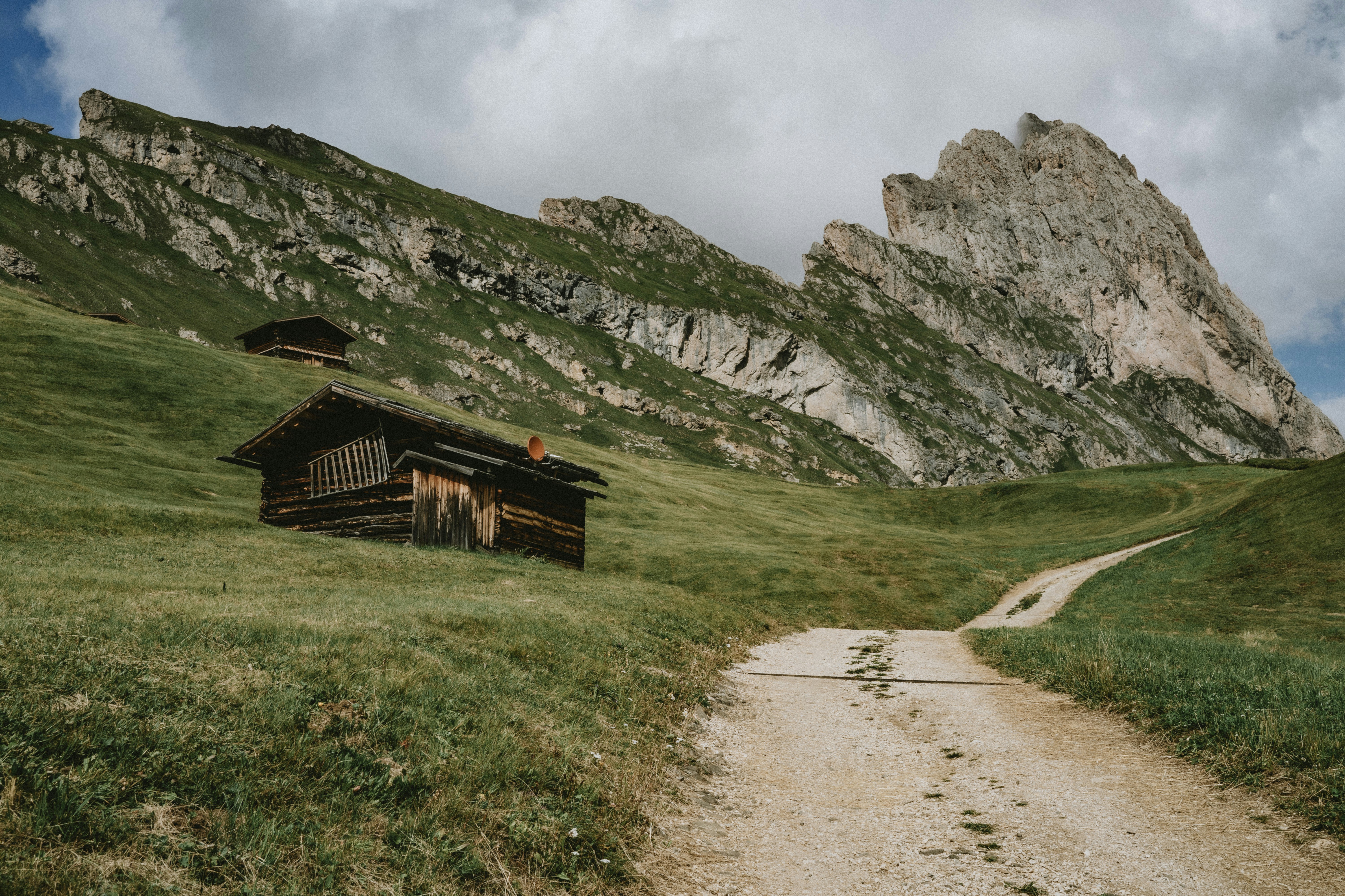 Rustic wooden cabins nestled in a lush green valley beneath towering mountains, with a winding path leading through the landscape.