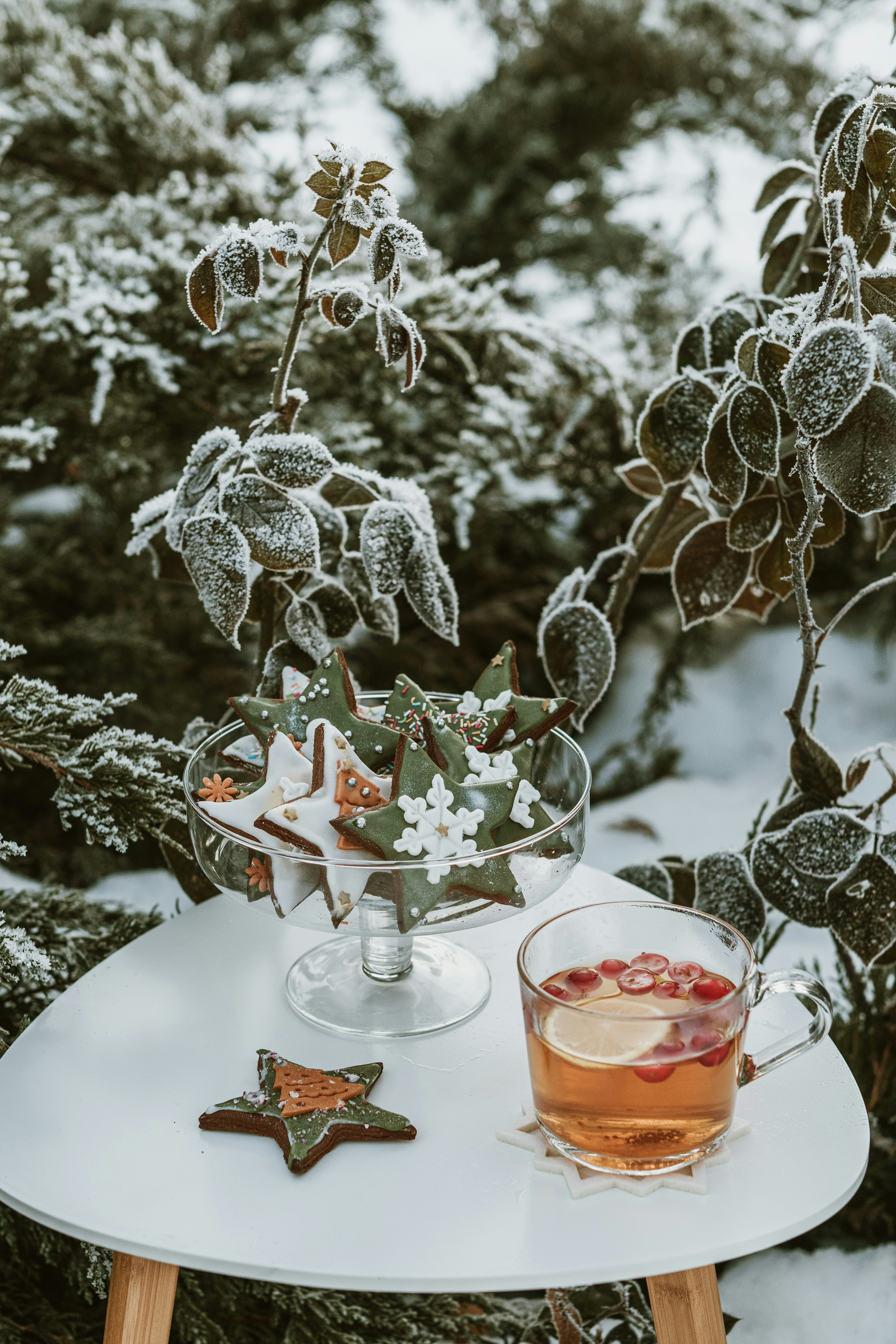 A glass dish filled with decorated star-shaped cookies sits on a white table, accompanied by a glass of tea adorned with floating berries, surrounded by frosted greenery.