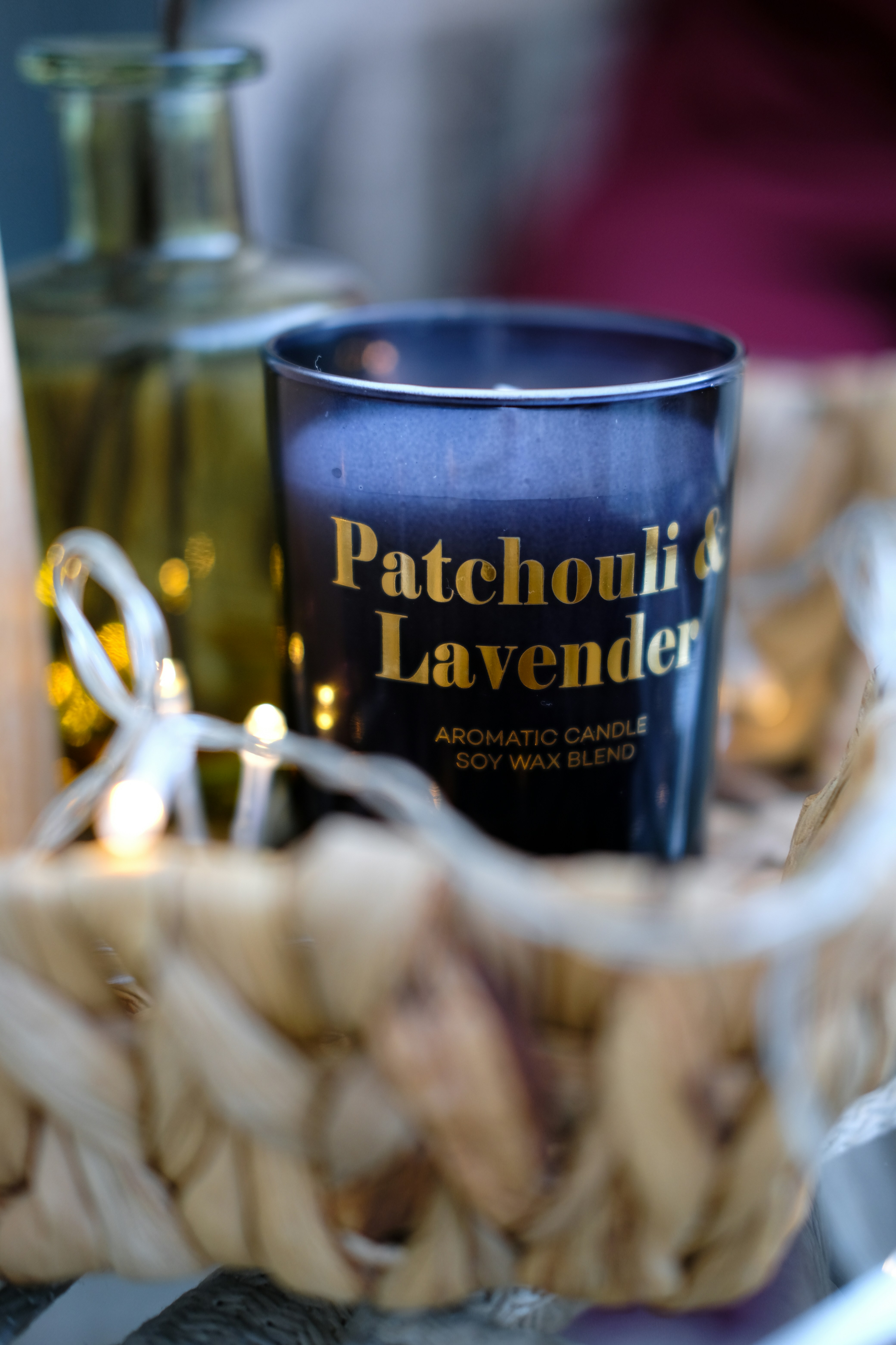 Smoky blue candle with Patchouli & Lavender labeling sits inside a woven basket, lit string lights and a green bottle in the background add warm ambience.