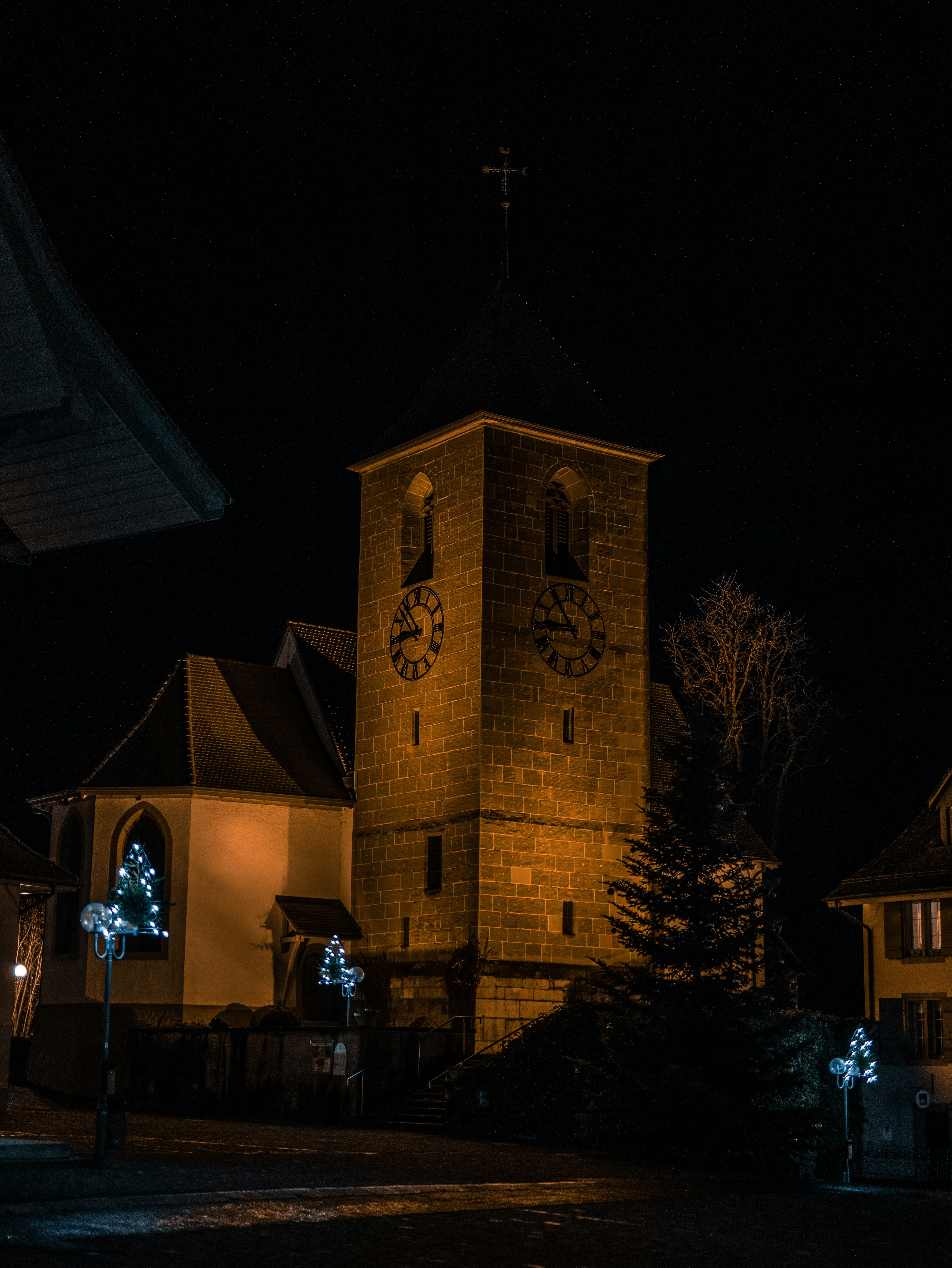 Historic clock tower illuminated against a dark sky, surrounded by festive lights and trees.