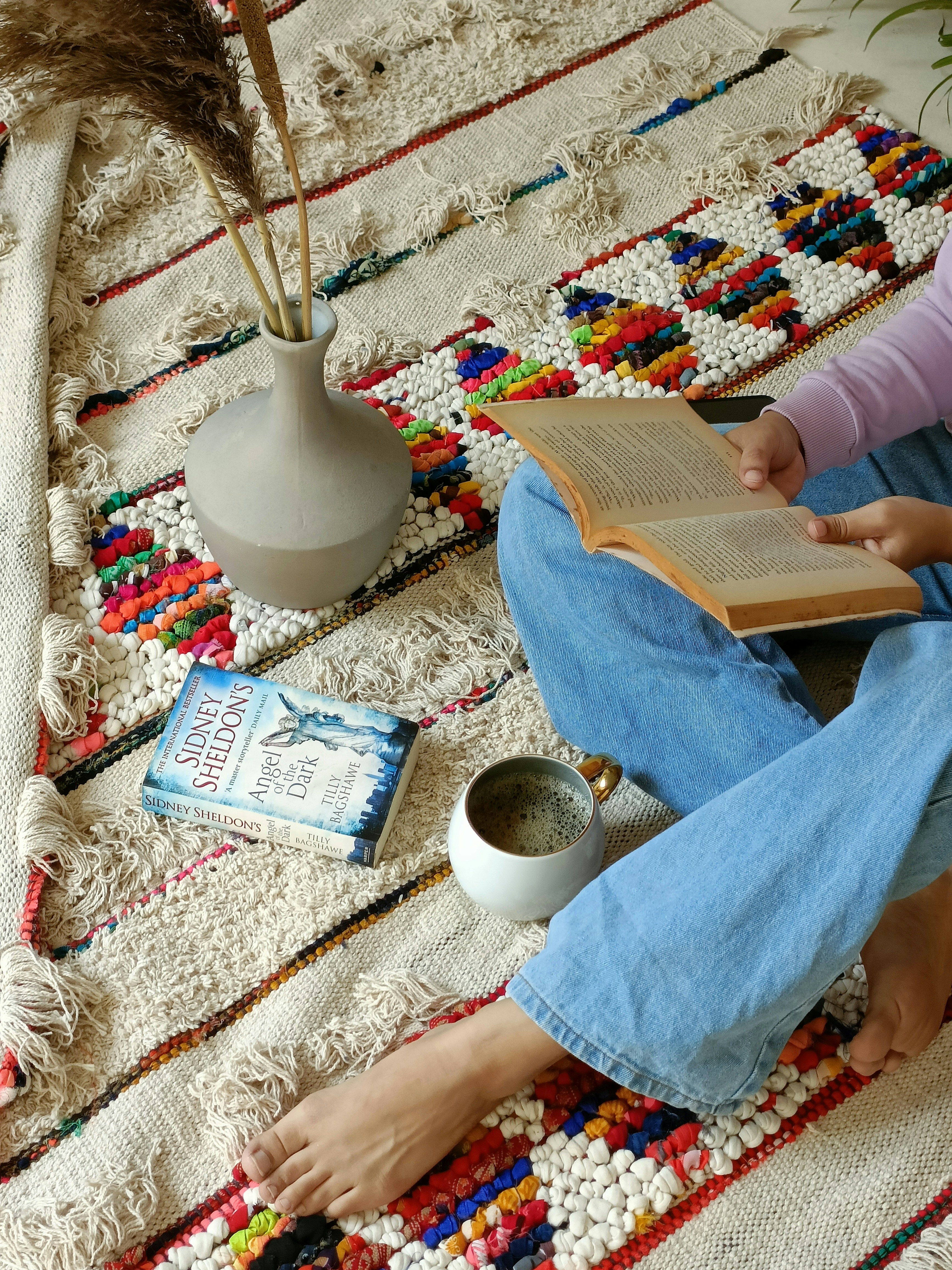 A person reading a book while seated on a colorful woven rug, with a coffee cup and a vase in the scene.