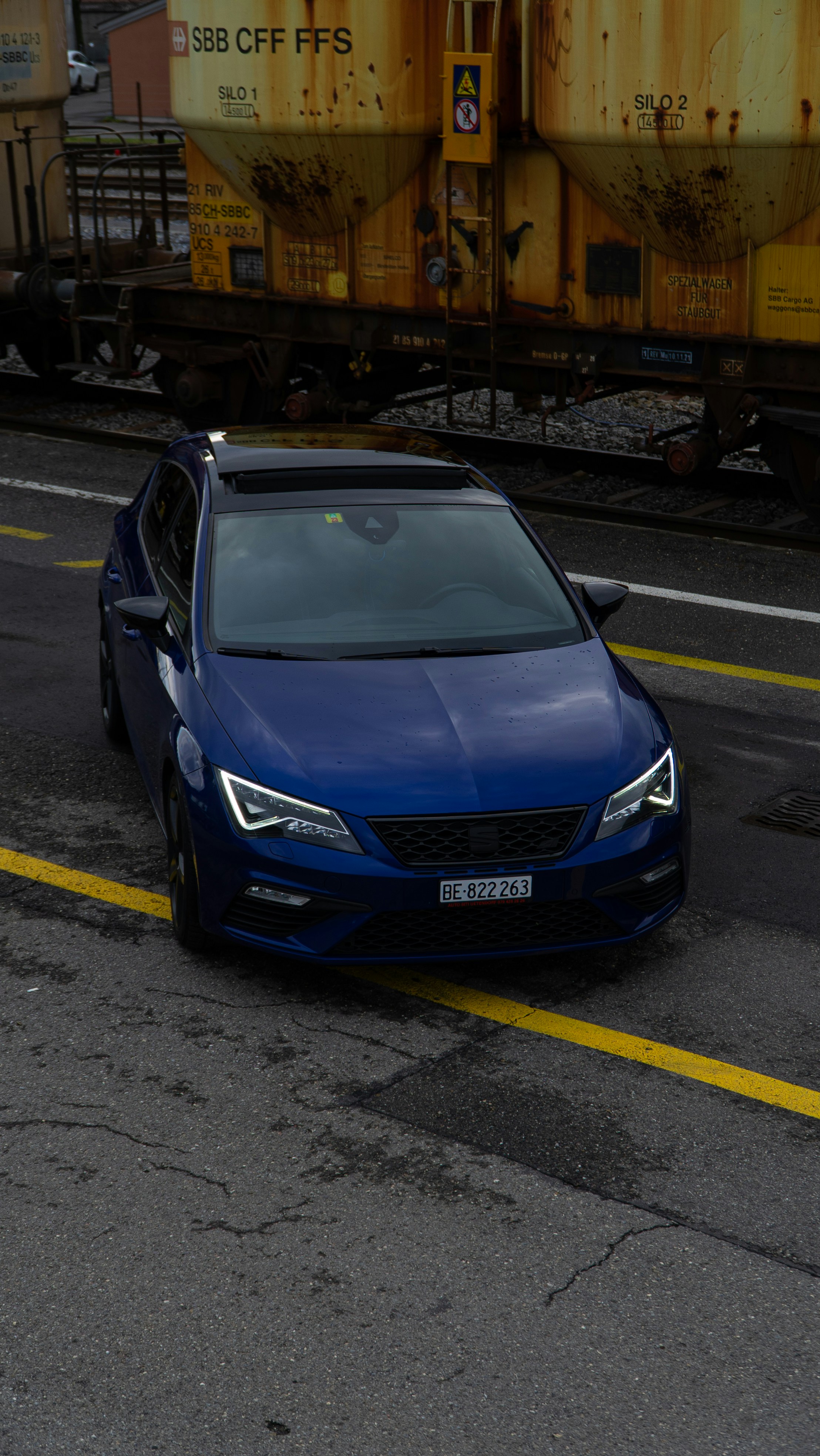 Blue sedan amid yellow freight giants: a quiet rail-yard portrait