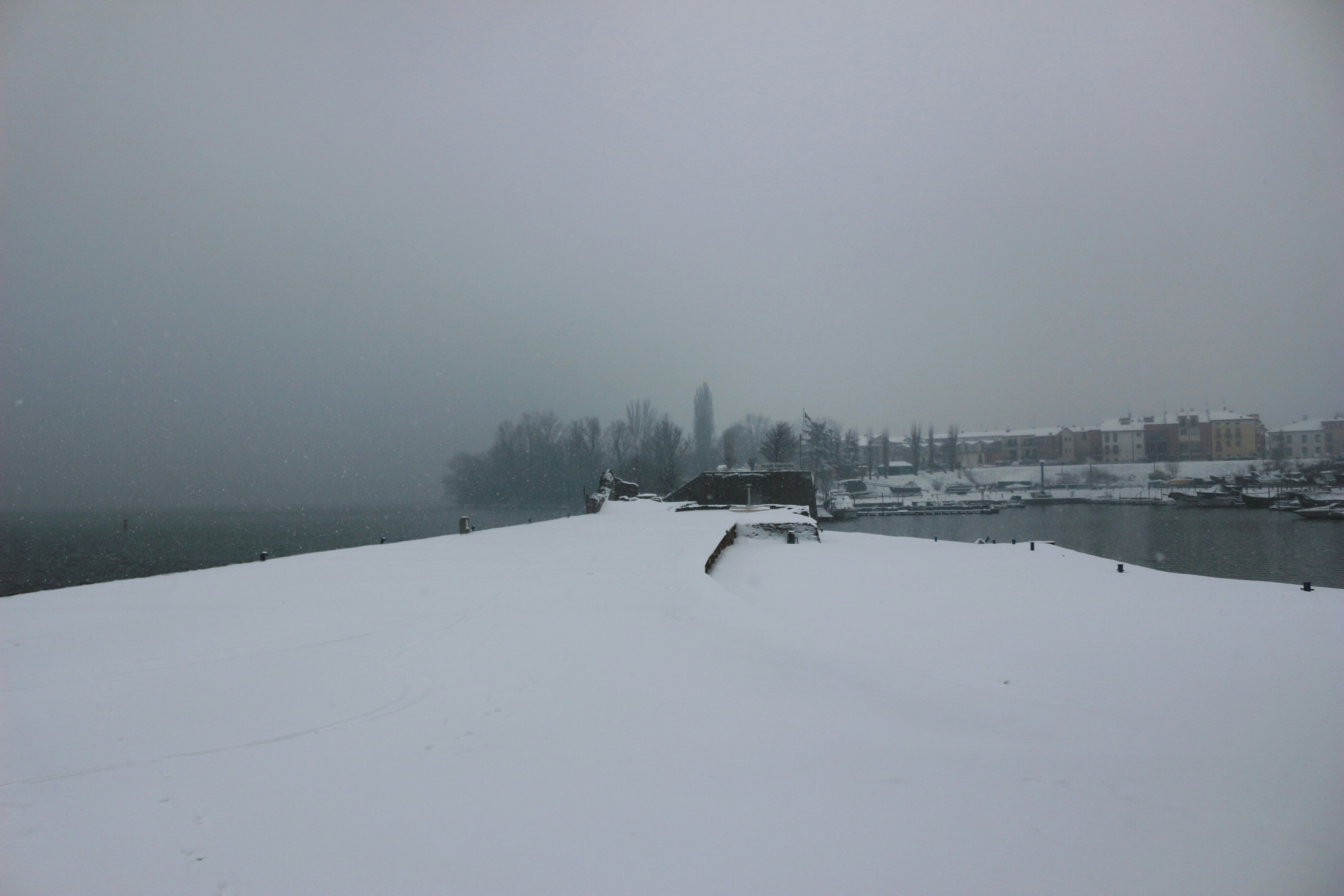 Snow-covered hill overlooking a misty body of water with distant trees and buildings.