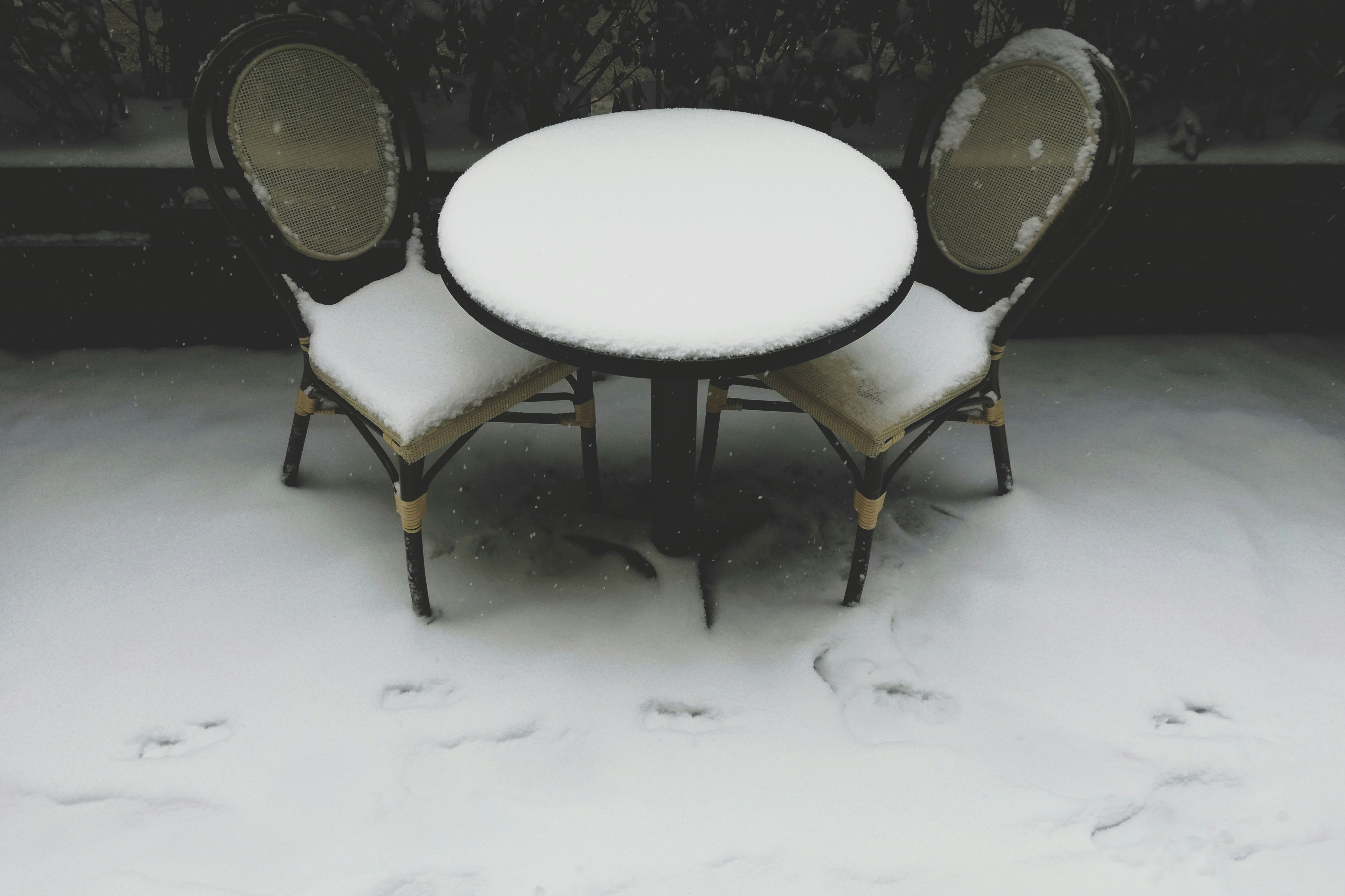 A round table and two chairs blanketed in snow, surrounded by a serene winter landscape. Footprints lead toward the scene, hinting at recent activity.