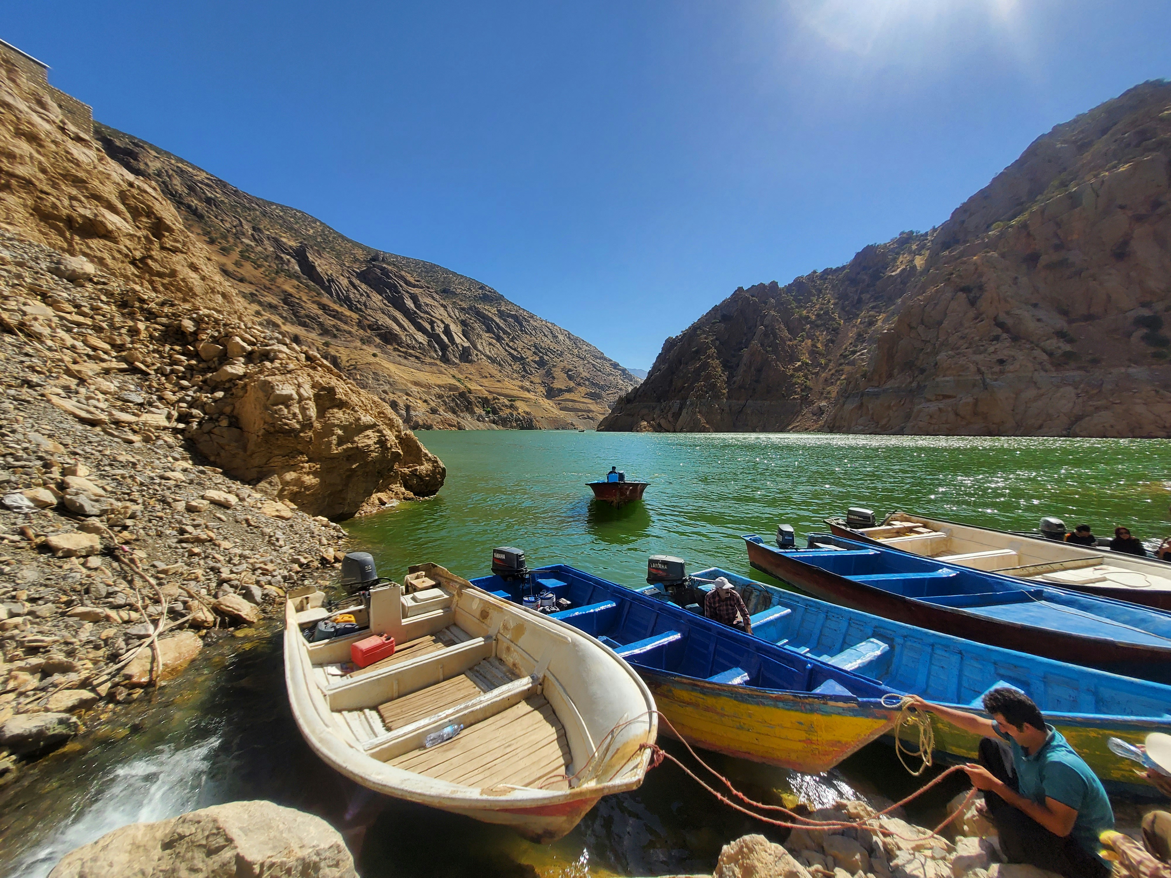 Small boats docked on a clear lake surrounded by rocky mountains under a bright blue sky.