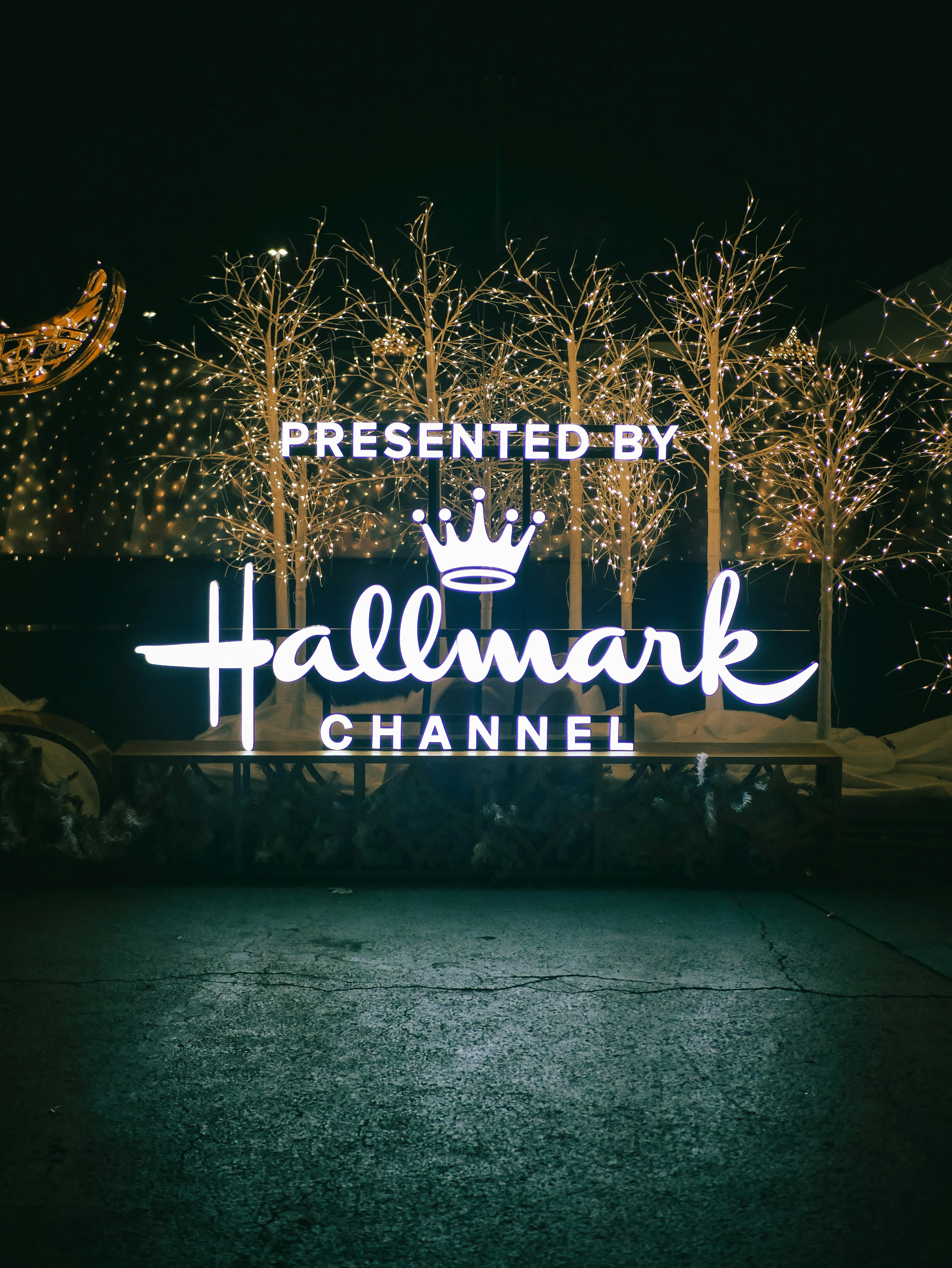 Neon sign featuring 'Presented by Hallmark Channel' against a backdrop of twinkling trees and festive lights.