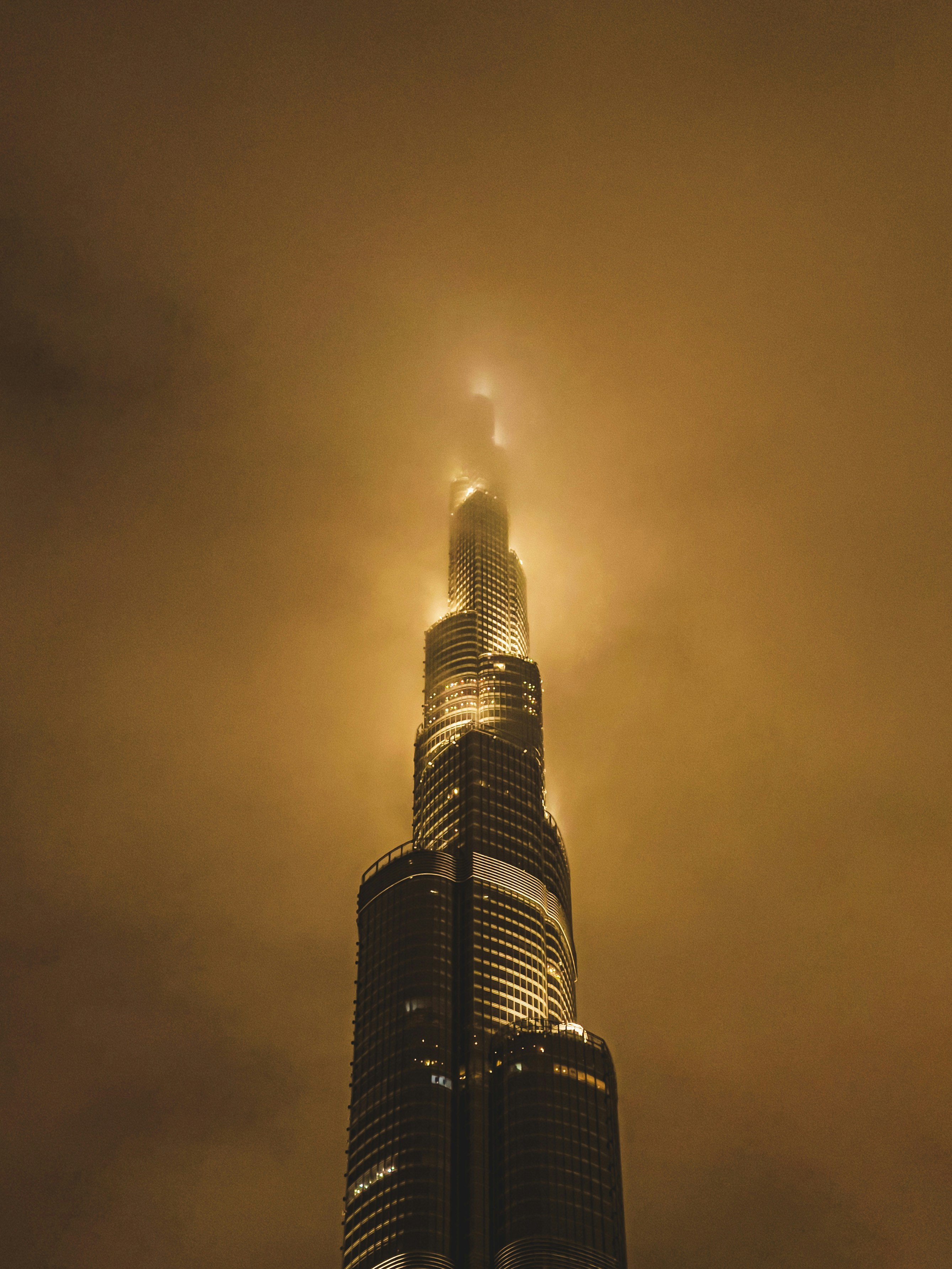 Burj Khalifa rises through dense fog bathed in an amber night glow, its illuminated tiers fading into the clouds.