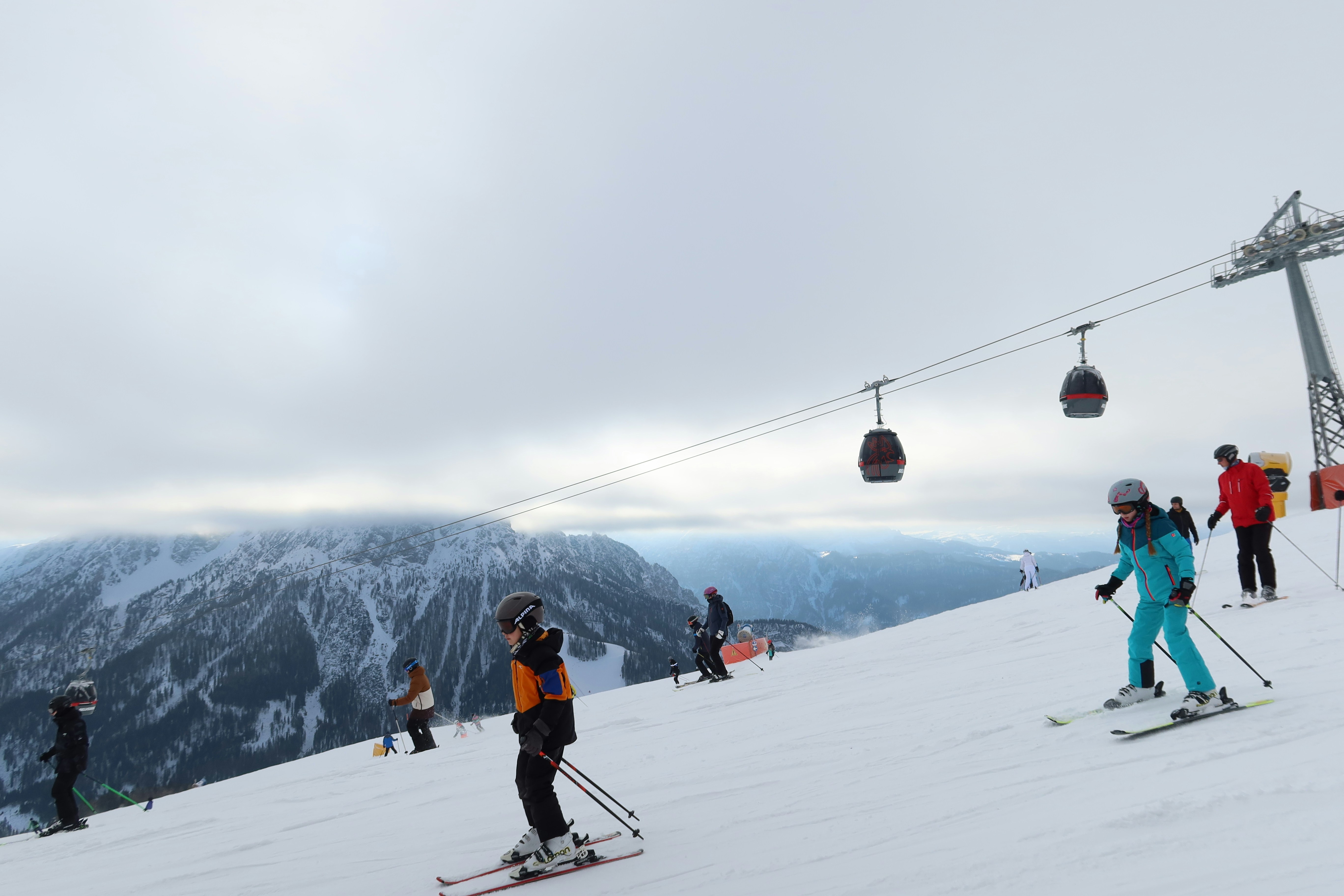 Skiers navigate snowy slopes beneath a cloudy sky, with distant mountains and cable cars in the background.