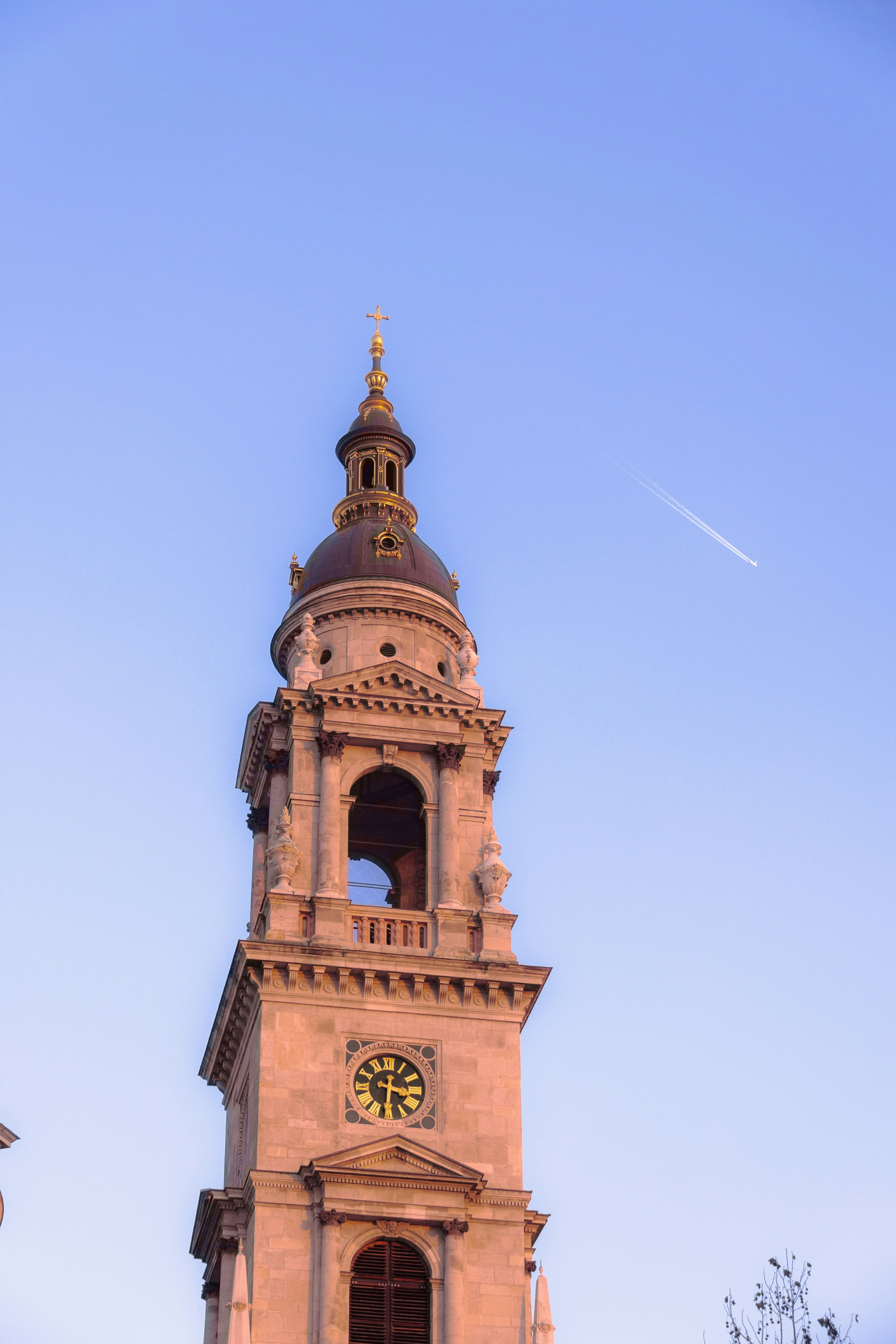 Historic clock tower reaching towards the evening sky, adorned with intricate details and a golden cross atop. A faint contrail hints at the passage of modernity above.