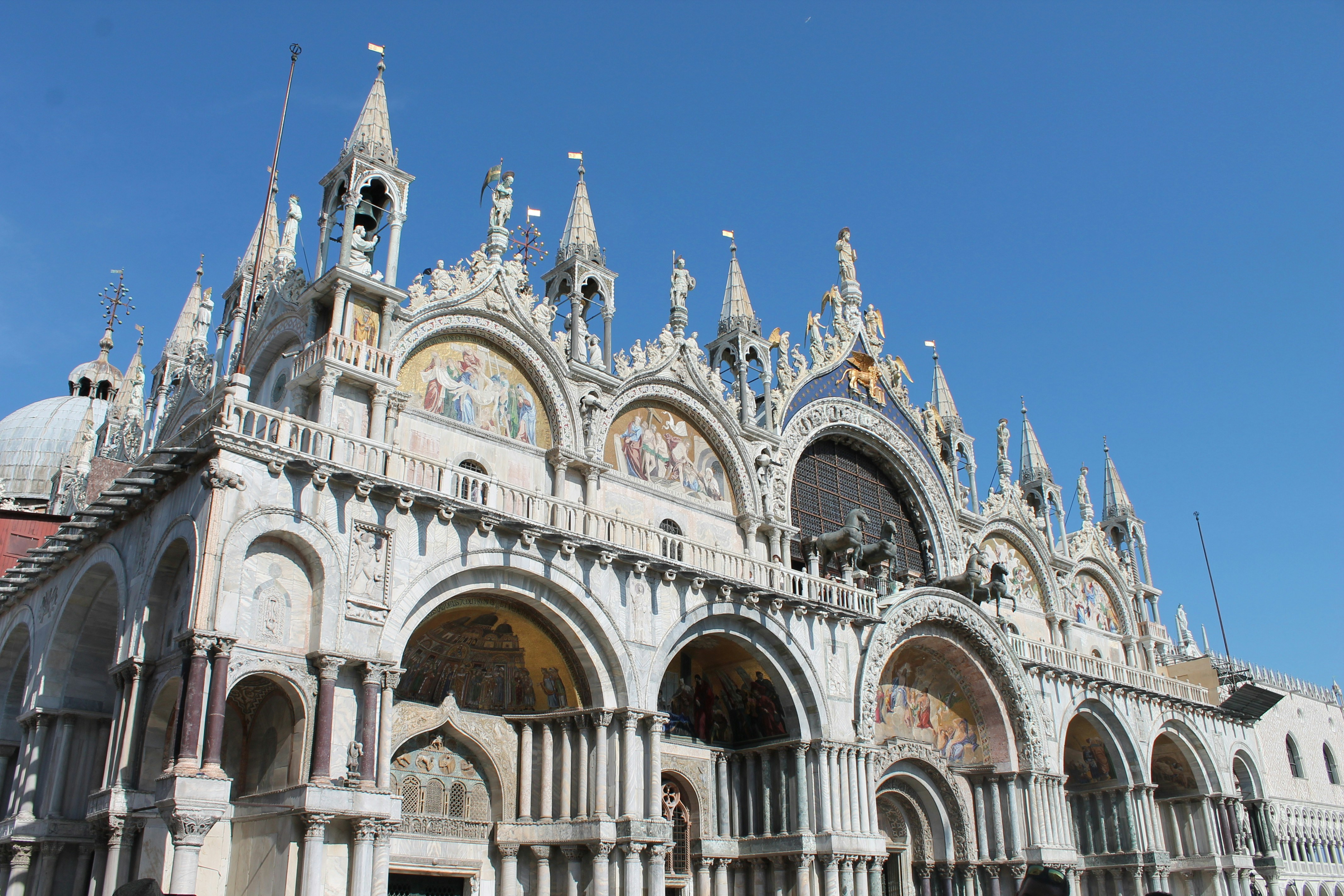 This image captures the intricate facade of St. Mark's Basilica in Venice, showcasing its stunning architectural details against a vibrant blue sky. The composition highlights the ornate arches, stone carvings, and colorful mosaics that adorn the historic structure. The bright, natural lighting accentuates the basilica's grandeur, creating a captivating and uplifting atmosphere.