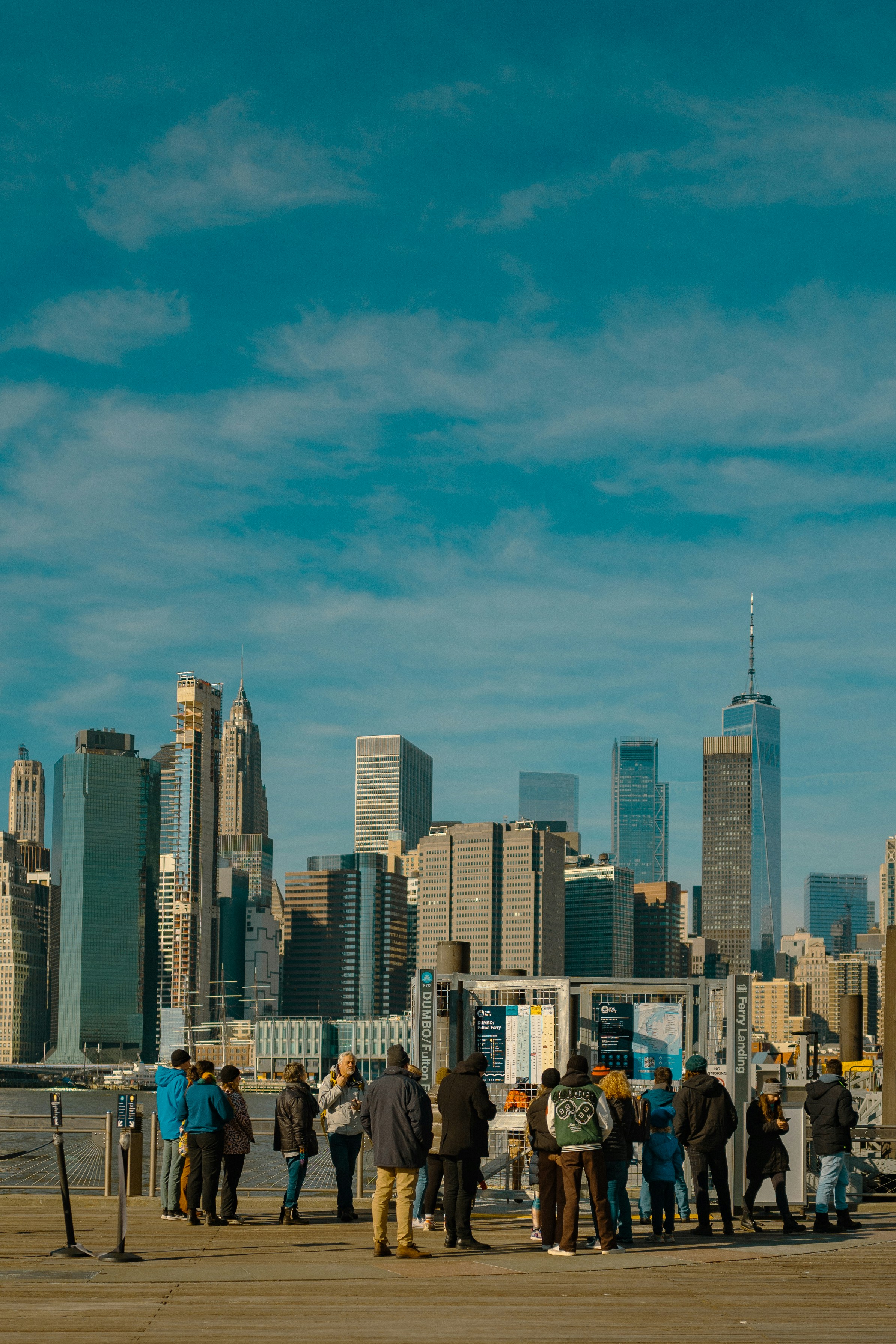 A diverse group of people gathered on a waterfront promenade, with a backdrop of towering skyscrapers under a clear blue sky.