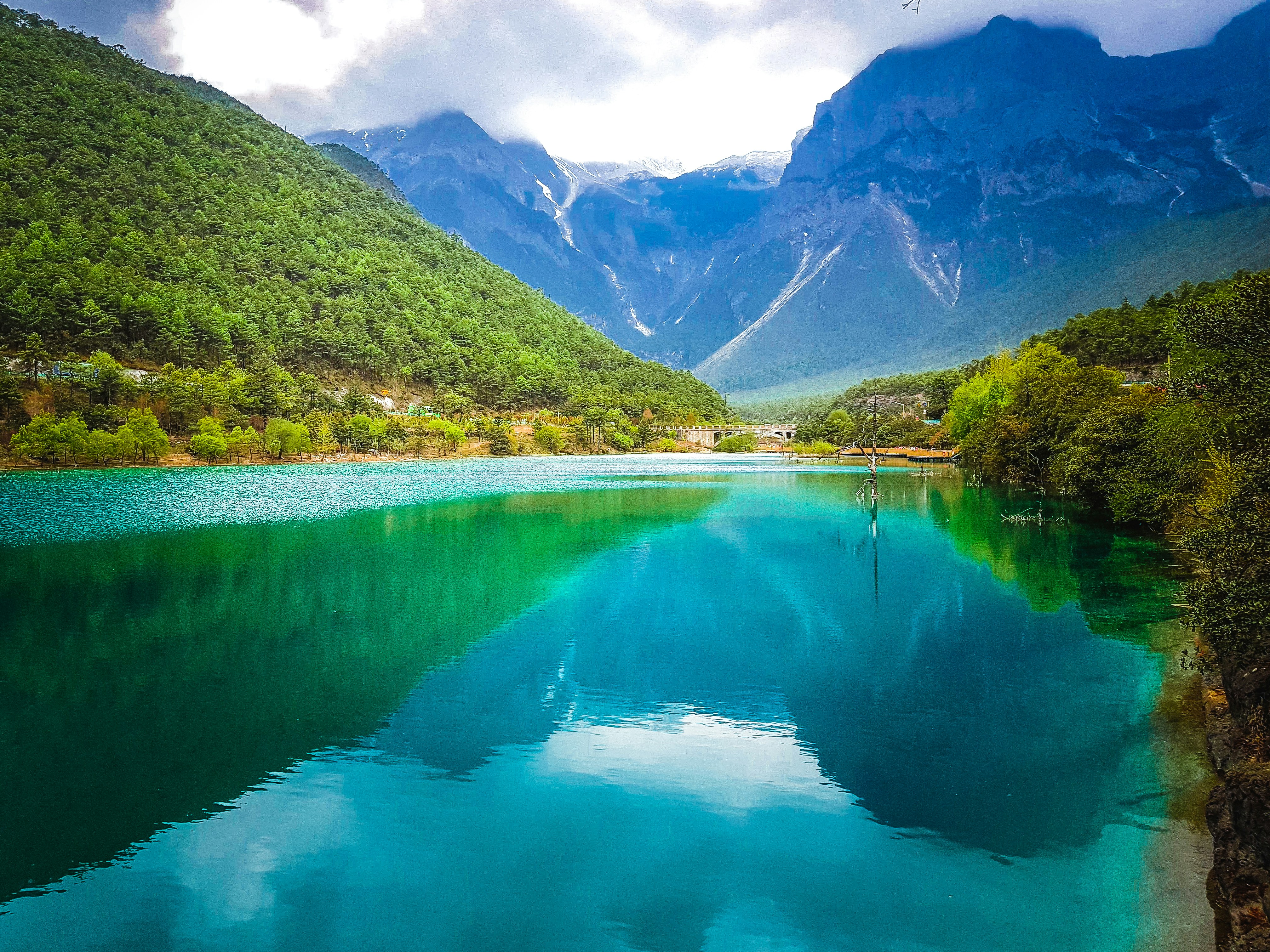 Turquoise lake reflecting majestic mountains and lush greenery under a cloudy sky. The tranquil scene invites contemplation.