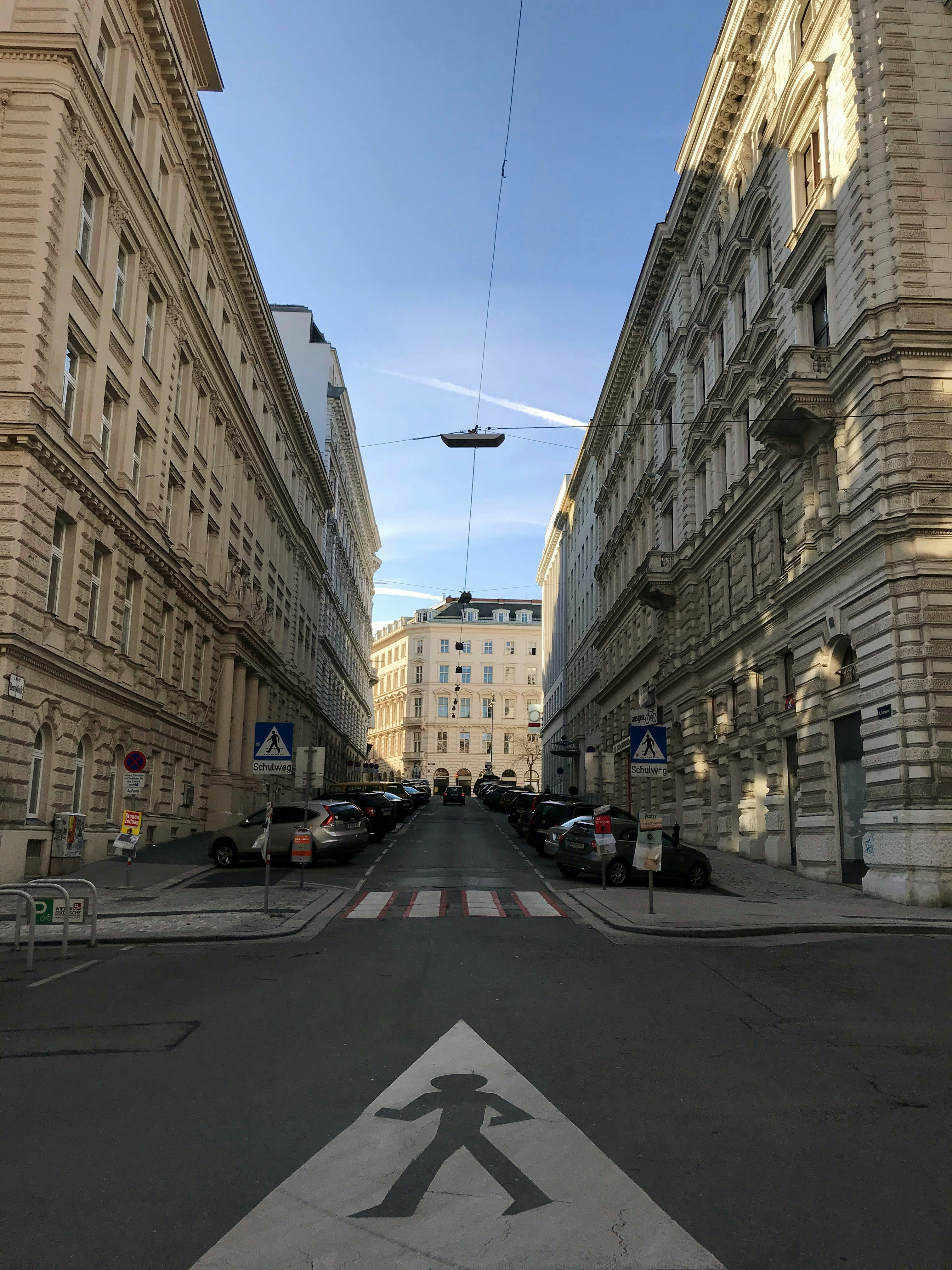Narrow street flanked by historic buildings, leading to a distant intersection with a clear blue sky above. Pedestrian crossing symbol prominently displayed on the road.