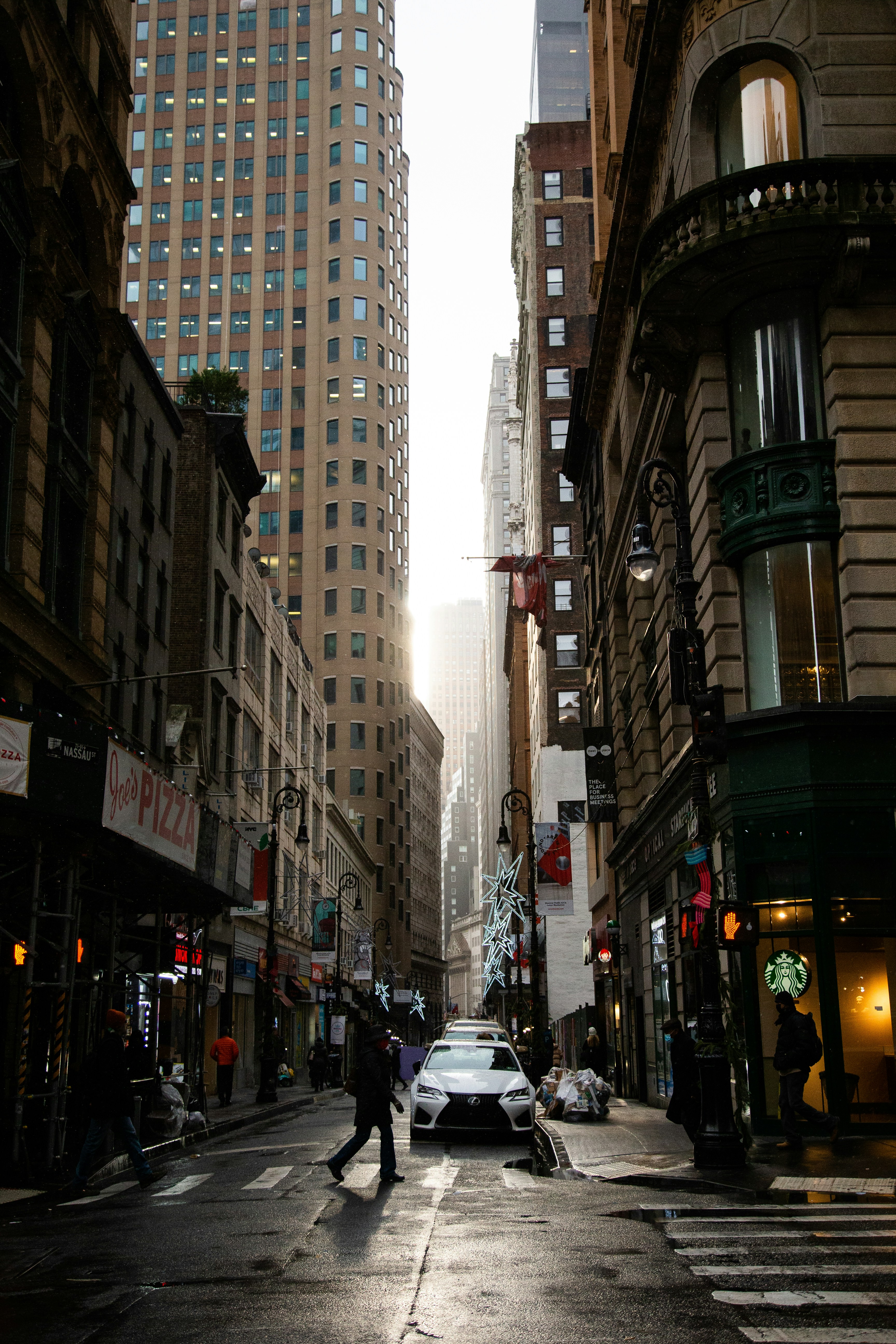 Narrow city street illuminated by sunlight breaking through towering skyscrapers, featuring pedestrians and storefronts adorned with seasonal decorations.
