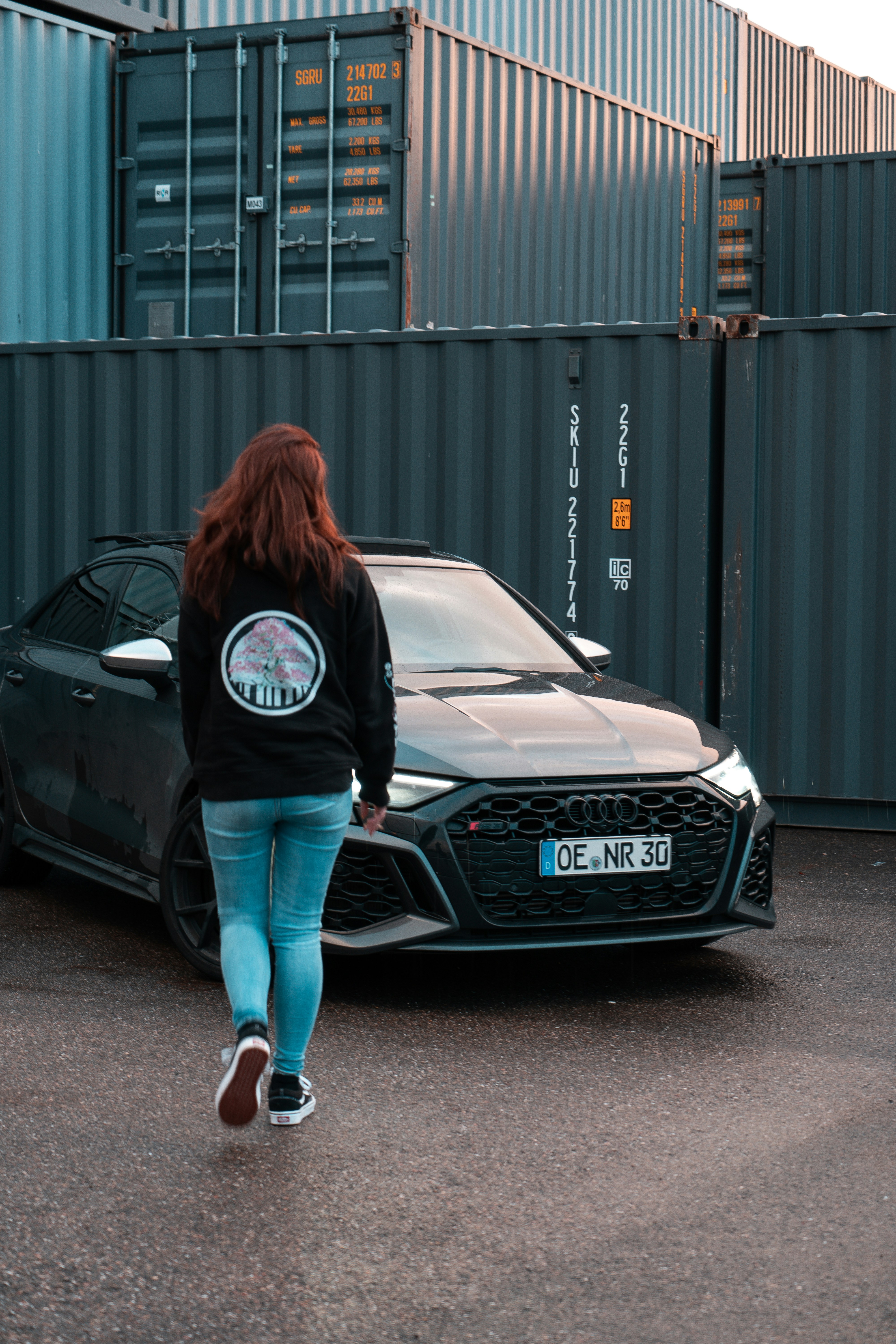 Red-Haired Walk Toward a Black Audi Amidst Container Towers