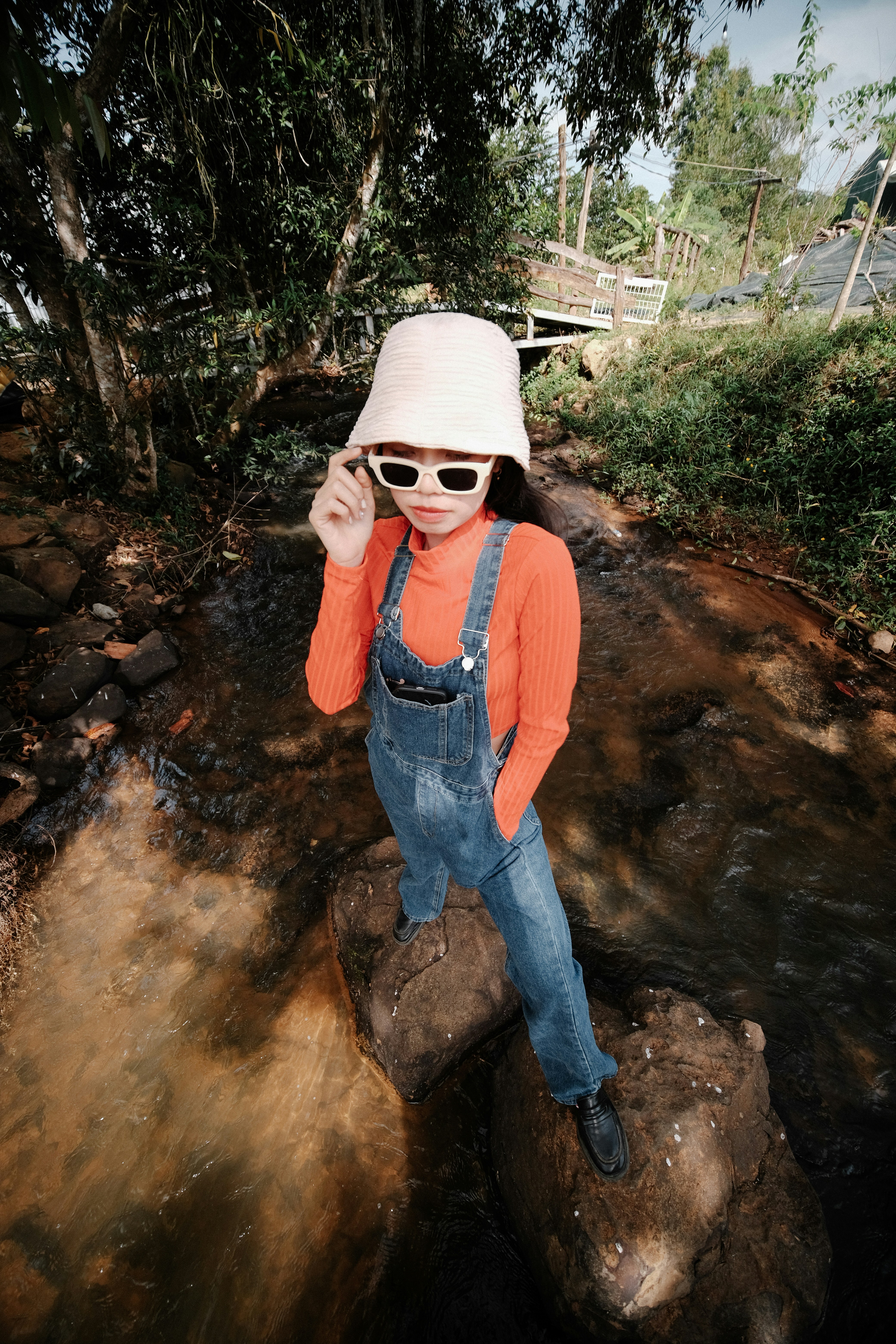 Girl in orange sweater and overalls perches on a rocky streambank wearing a sunhat and white sunglasses