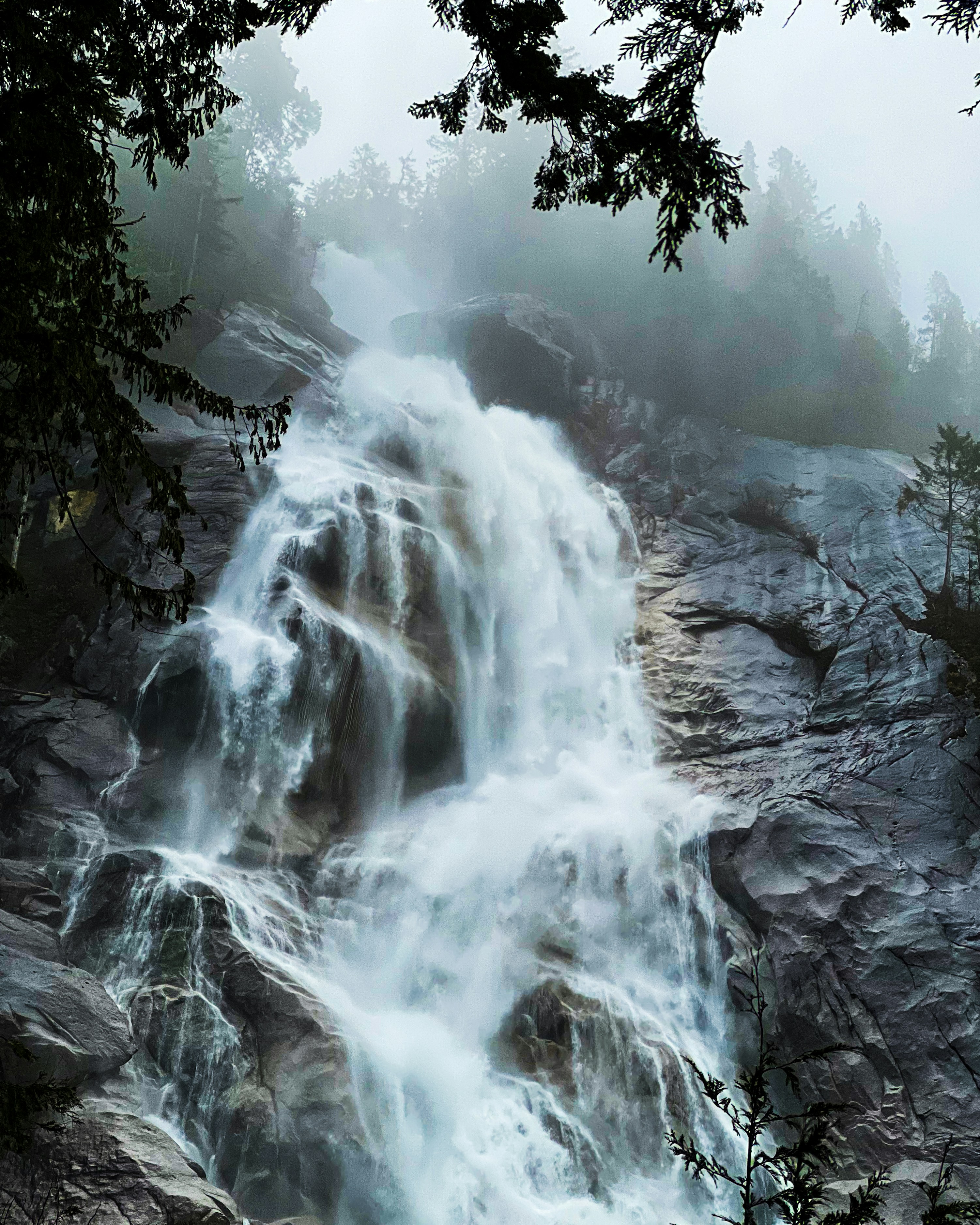 A majestic waterfall tumbles down rocky cliffs, enveloped in mist and framed by lush greenery. The scene evokes a sense of tranquility and natural beauty.