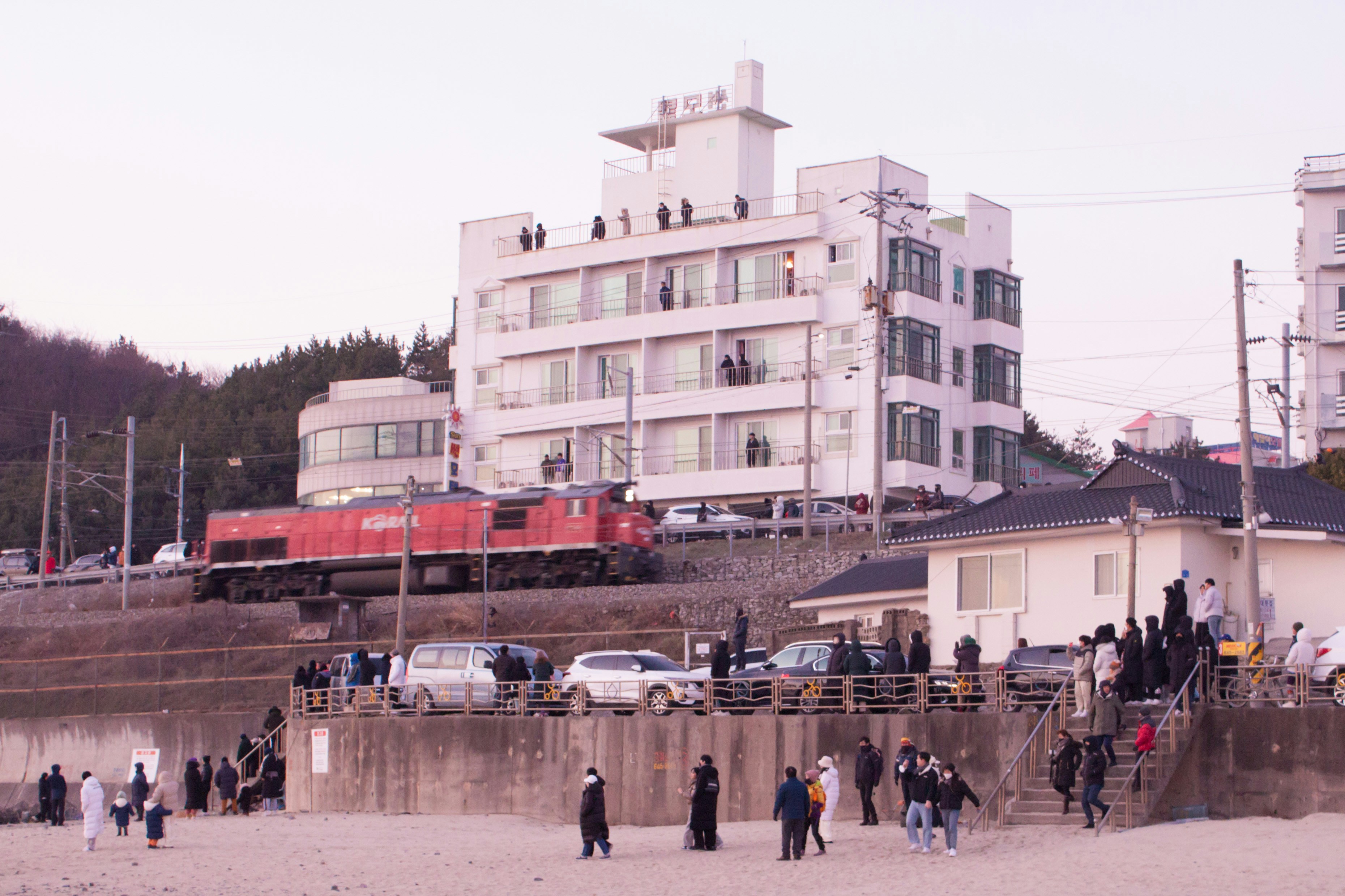 A red train speeds past a coastal building while crowds gather along the beach, capturing a moment of urban life meeting nature.