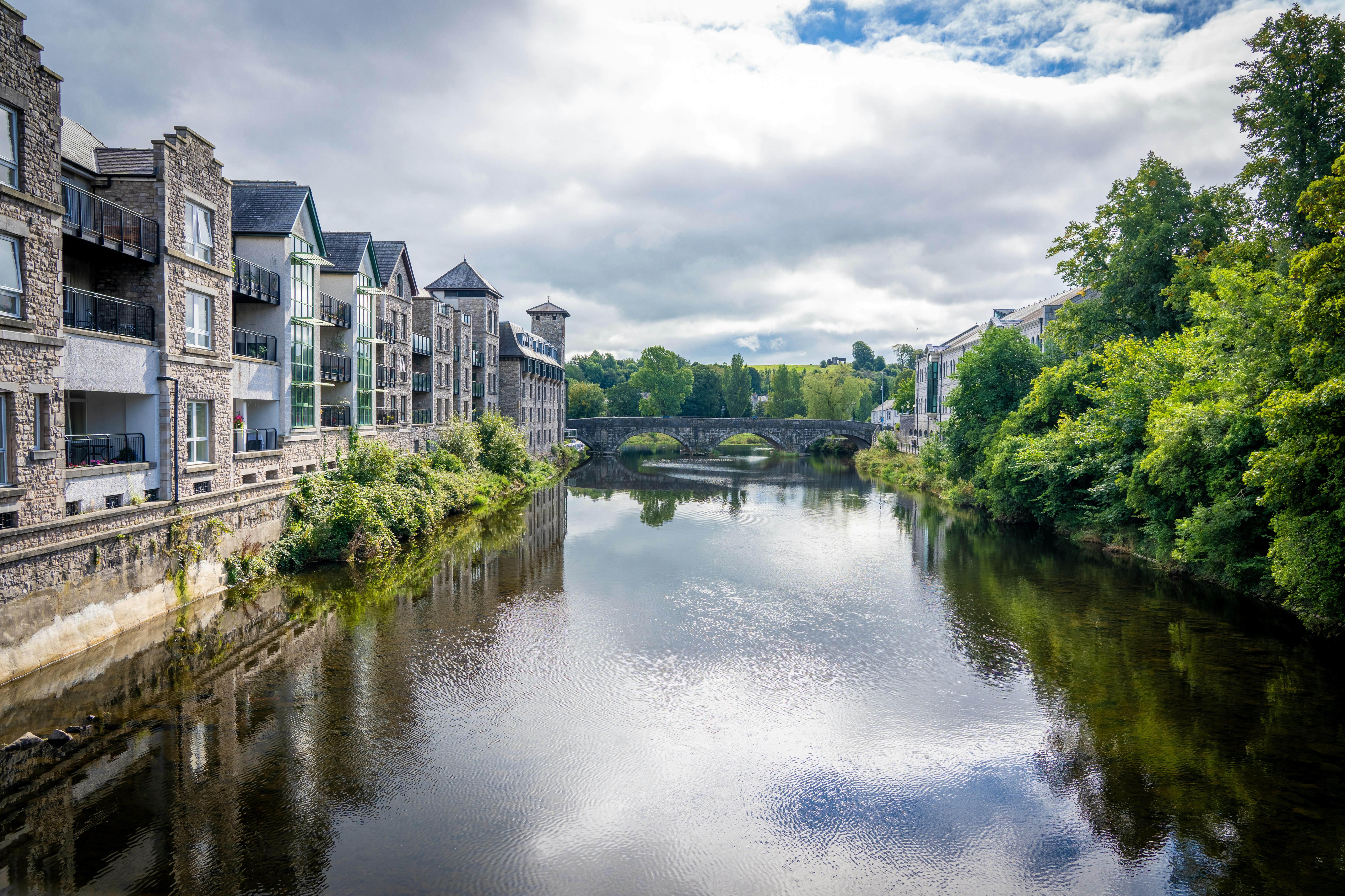 Tranquil river flowing between modern buildings and lush greenery under a cloud-dappled sky.