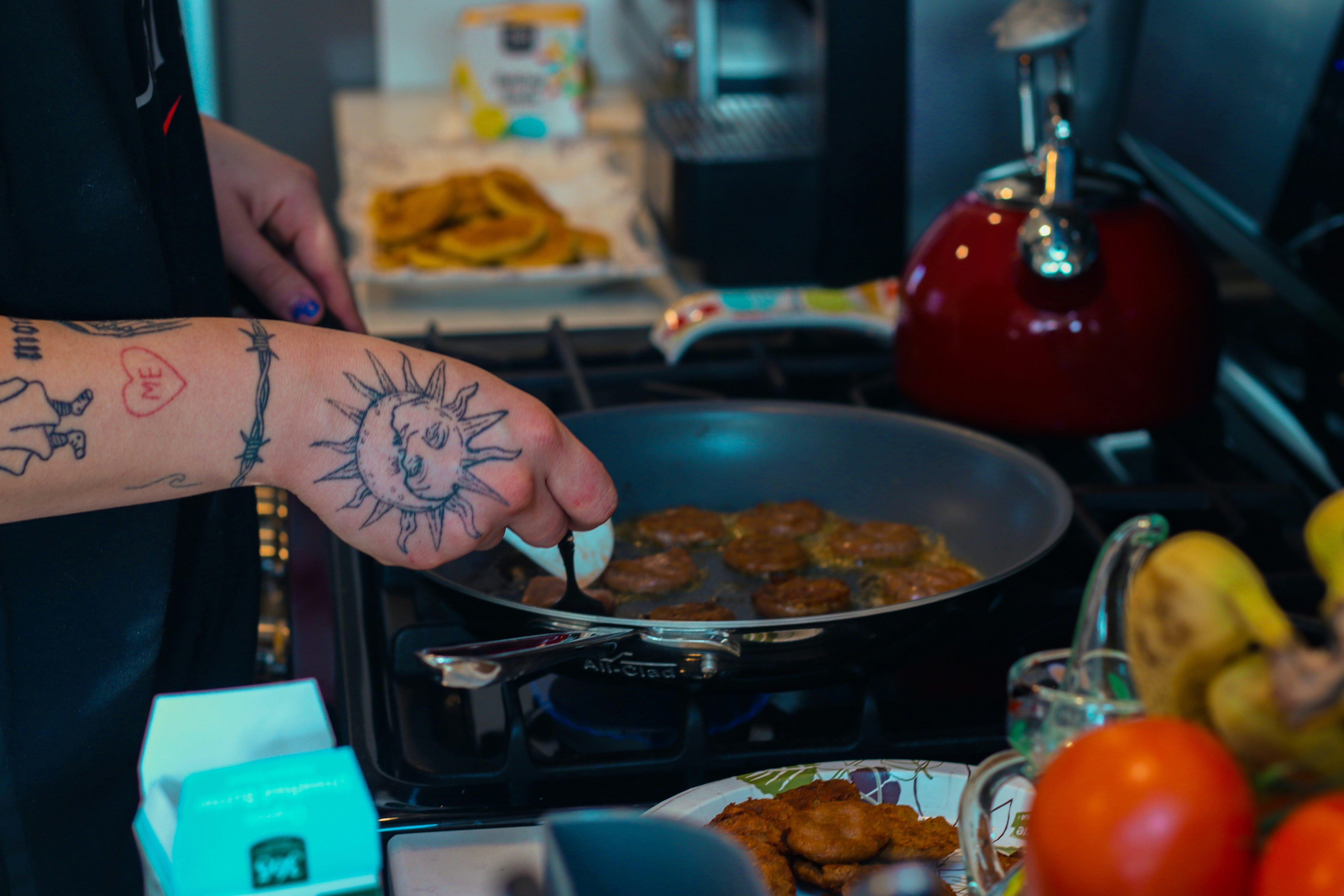 Sun-Tattooed Hand Navigates a Sizzling Skillet in a Blue-Hued Kitchen