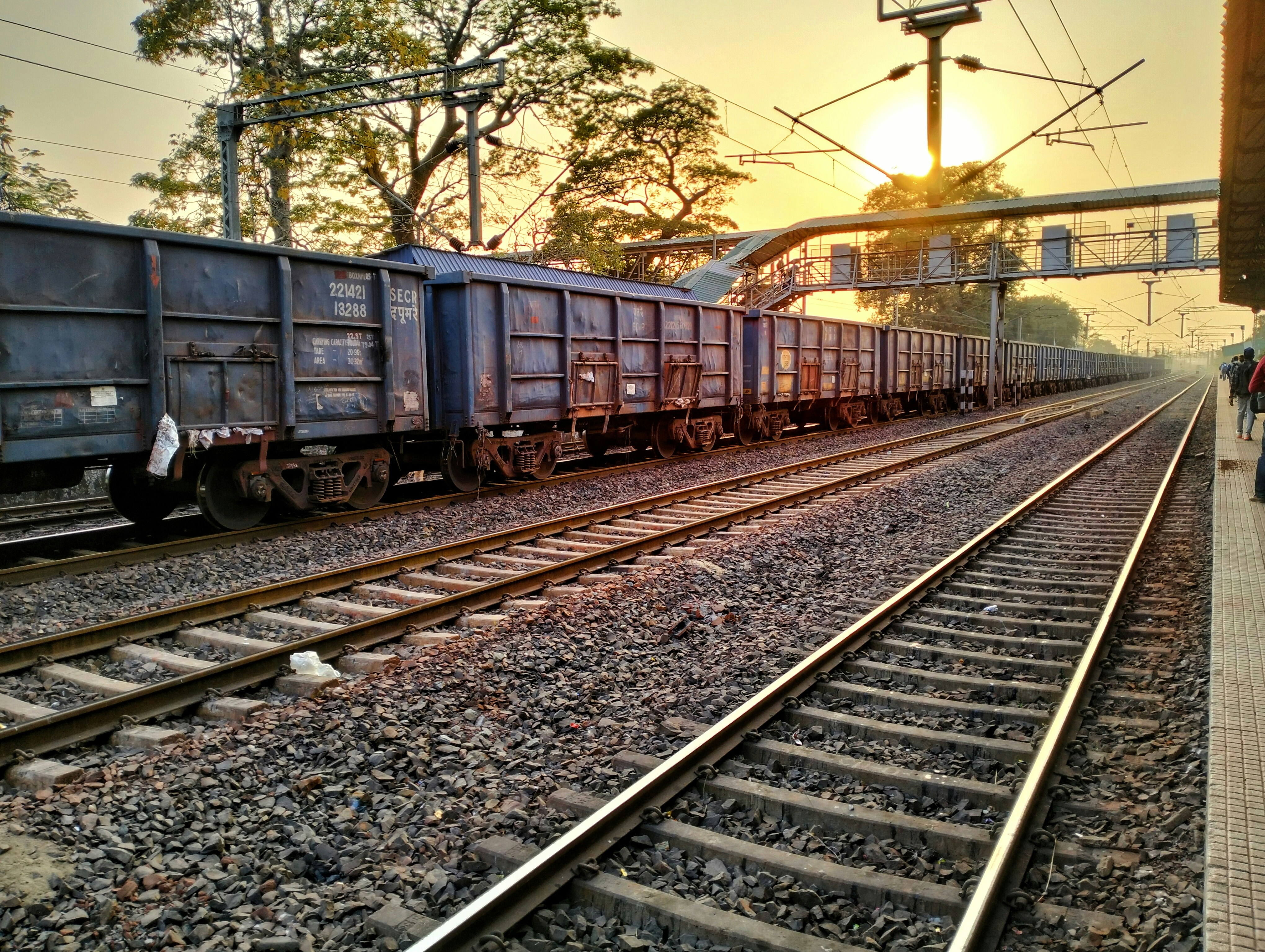Freight wagons line parallel tracks at sunset, bathed in warm light. Converging rails lead the eye toward a distant platform under a glowing sky.