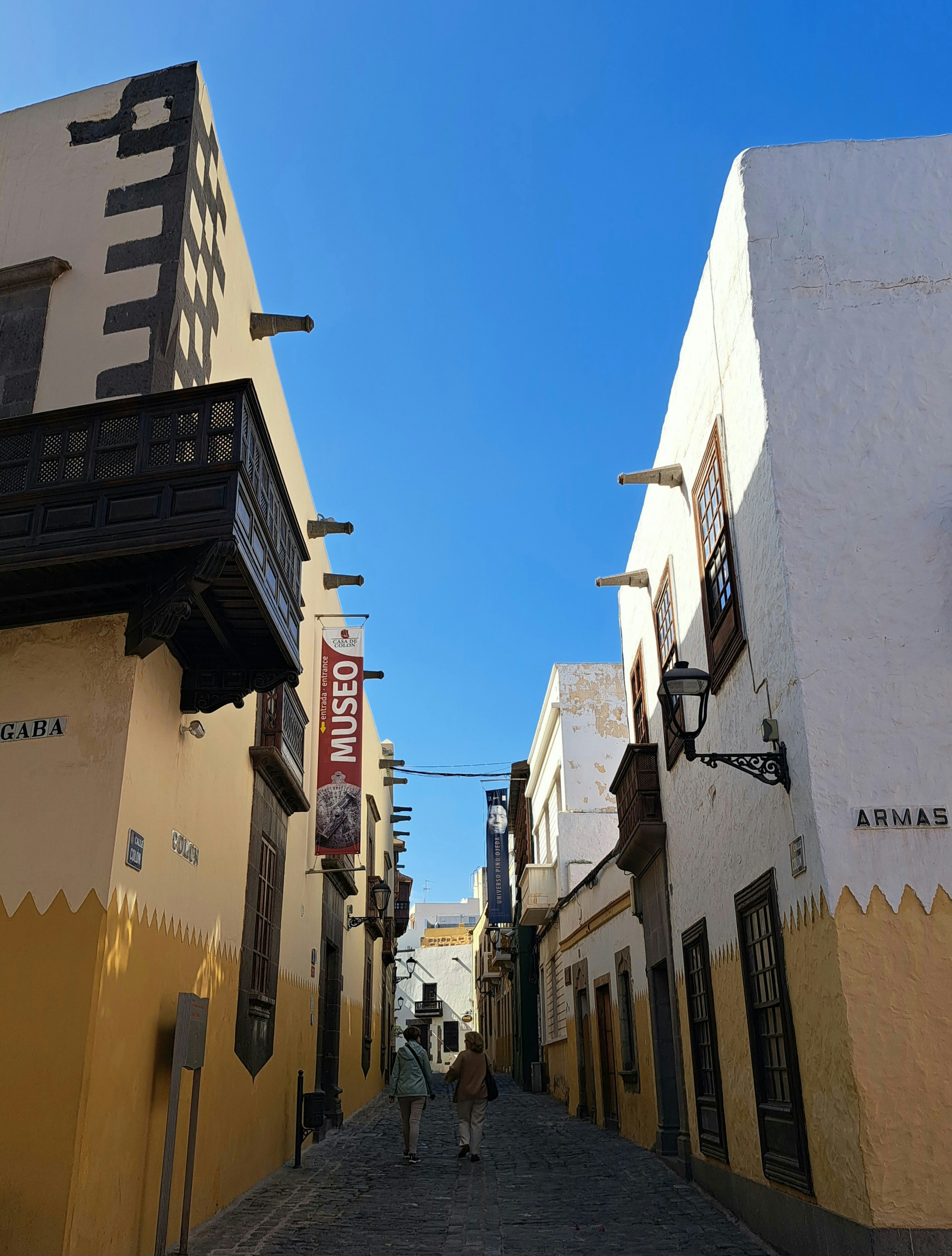 Charming narrow street flanked by traditional architecture, with a prominent museum sign and two figures walking towards the end. Bright blue sky overhead.