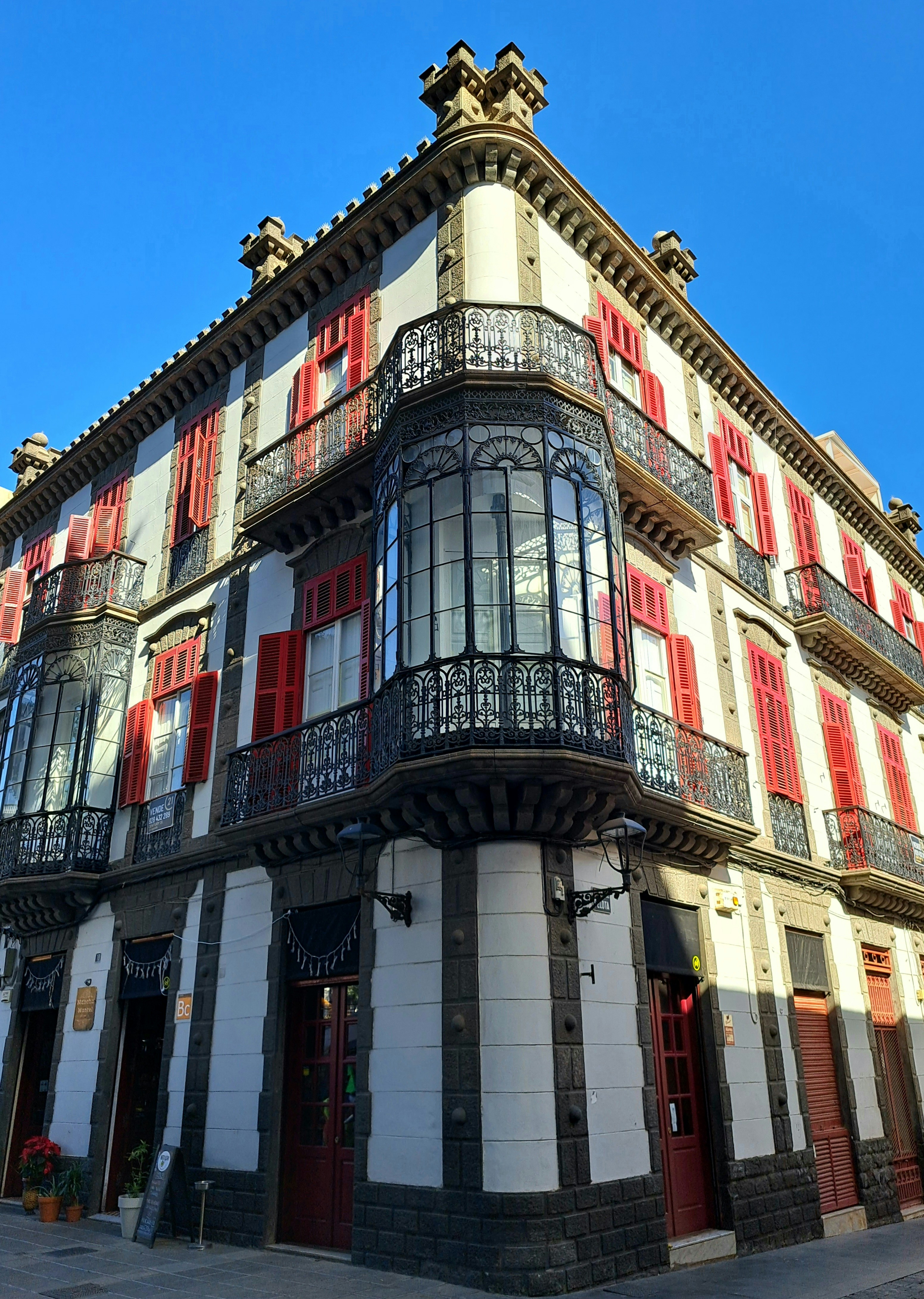 Artful building facade featuring intricate ironwork balconies and vibrant red shutters against a clear blue sky.