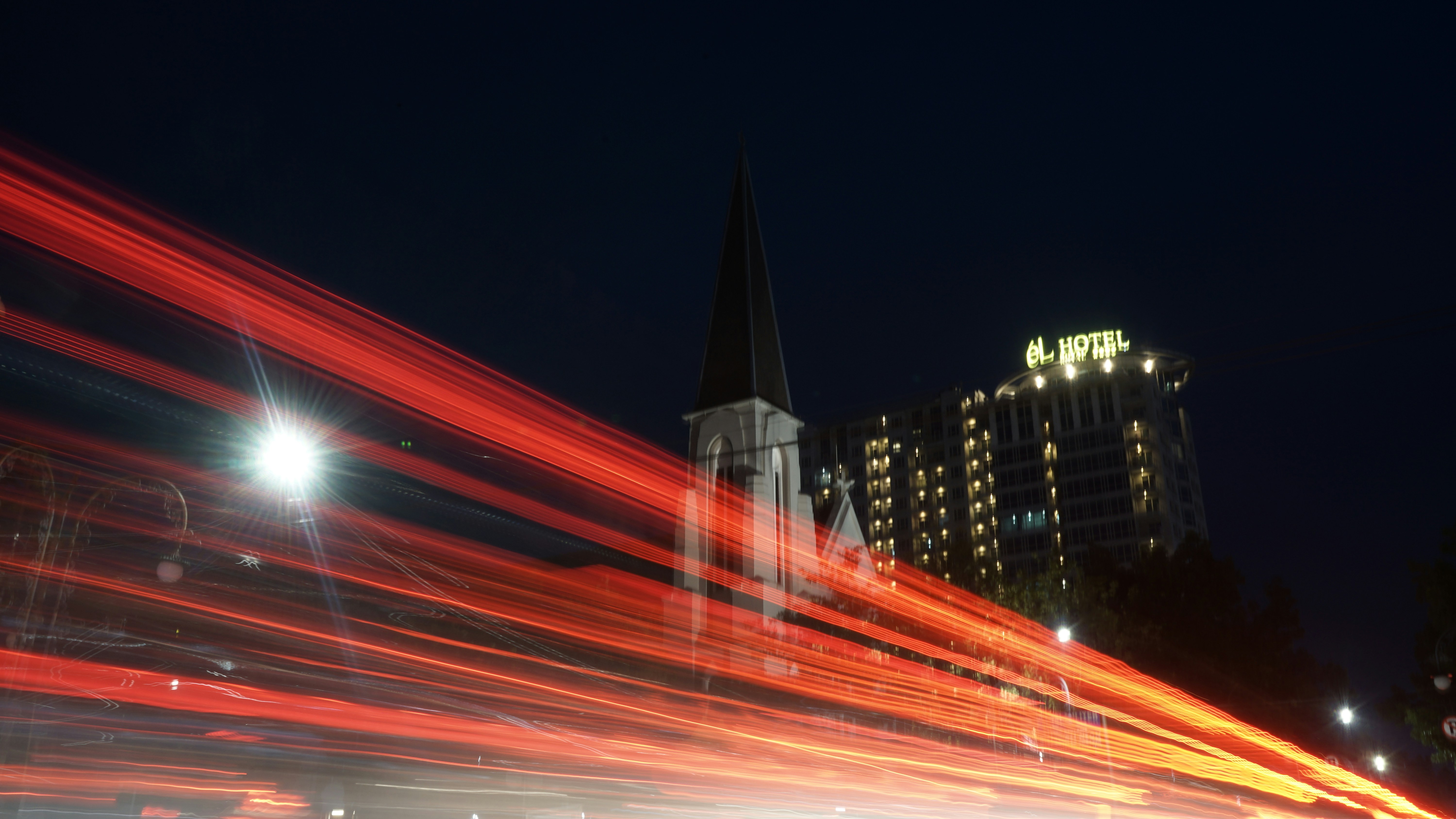 Dynamic light trails streak past a historic church, contrasting with a modern hotel backdrop at night.