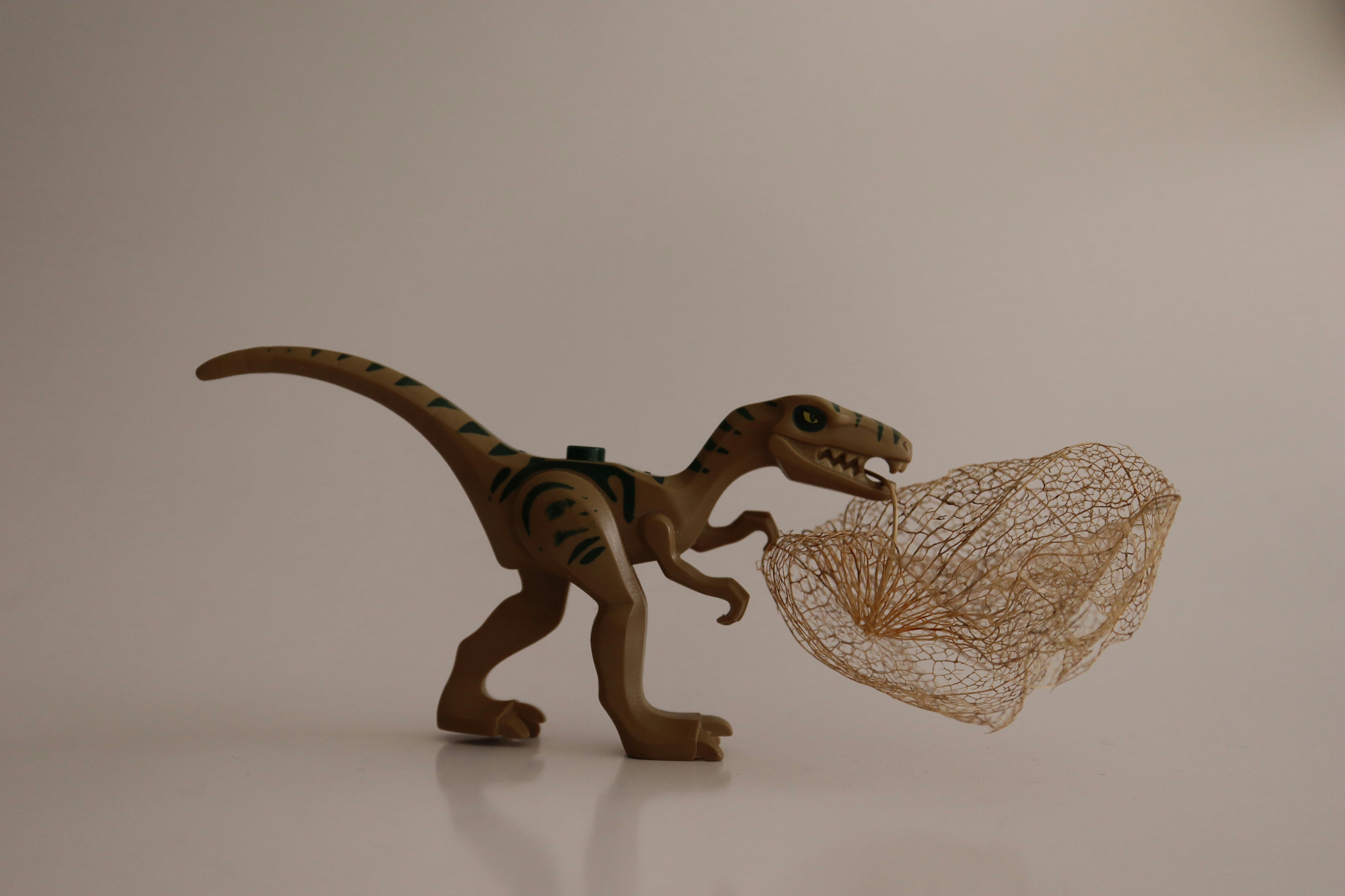 A toy dinosaur lunges forward, grasping a delicate net in its jaws, set against a soft, neutral background.